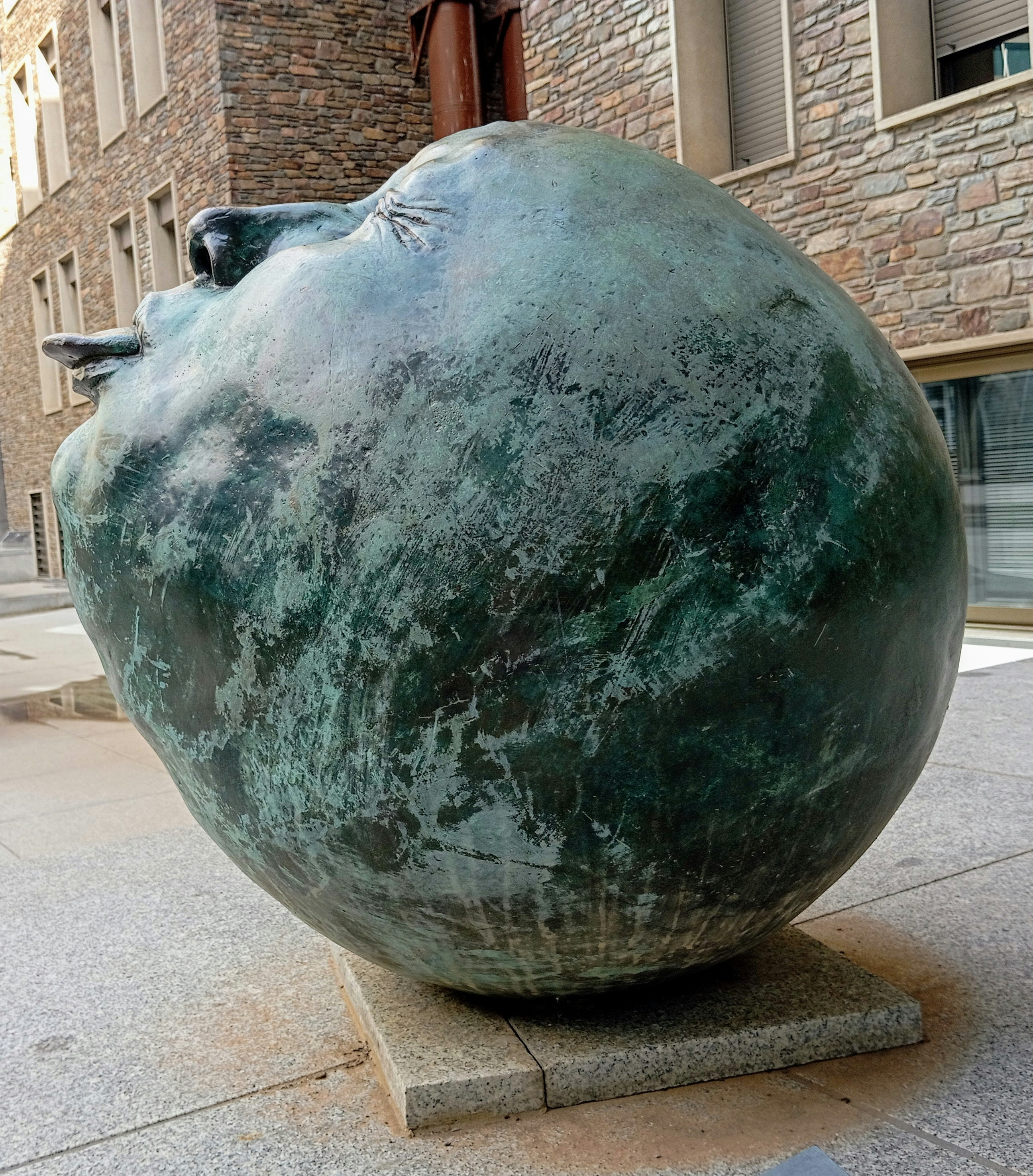 Verdigris bronze globe sculpture on a granite pedestal in an urban plaza, with a brick-and-stone building in the background.