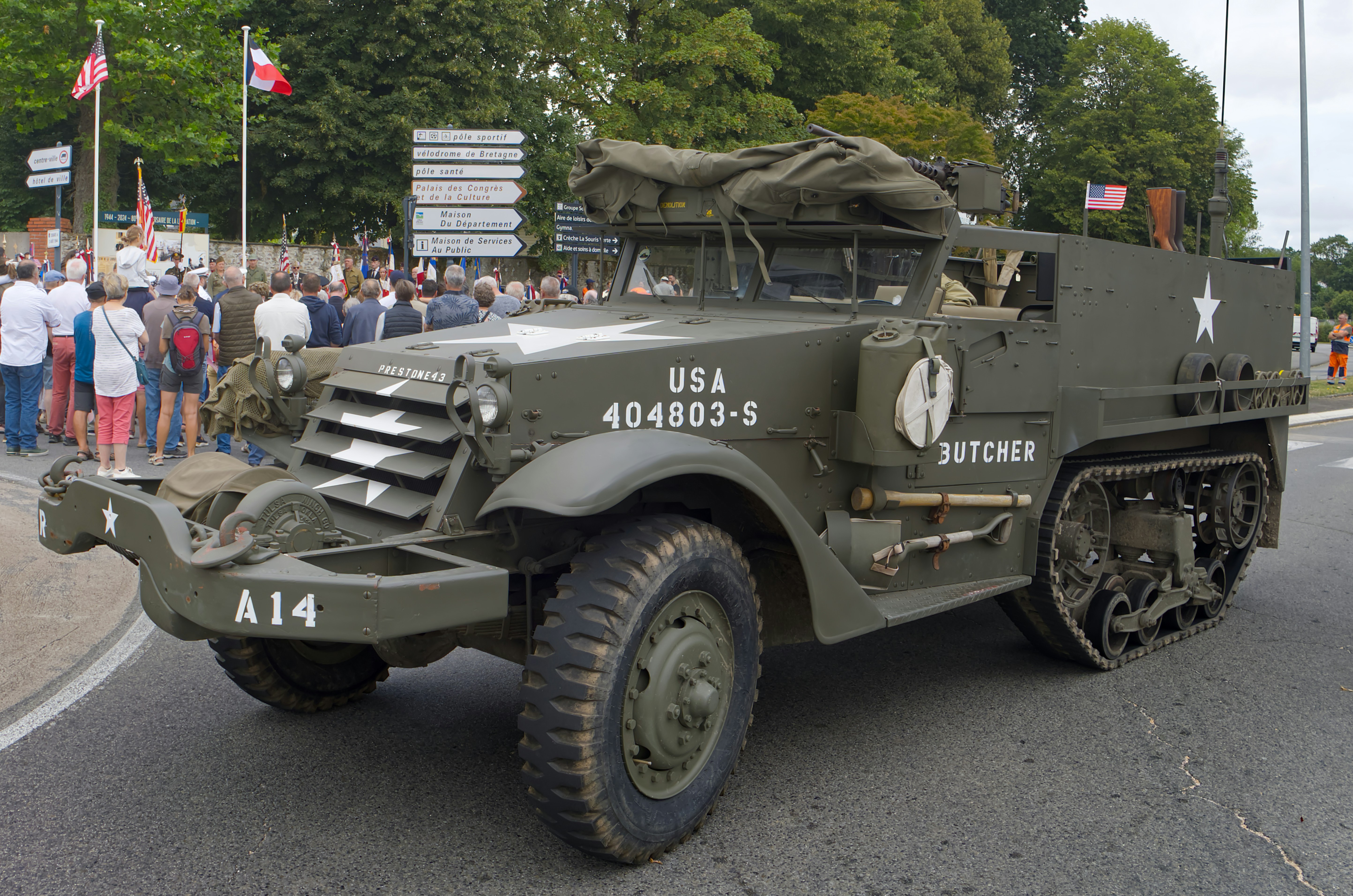 An historic US Army half-track rolls through a street parade, surrounded by spectators and waving flags. The armored vehicle bears stenciled markings and a white star, signaling its World War II heritage.