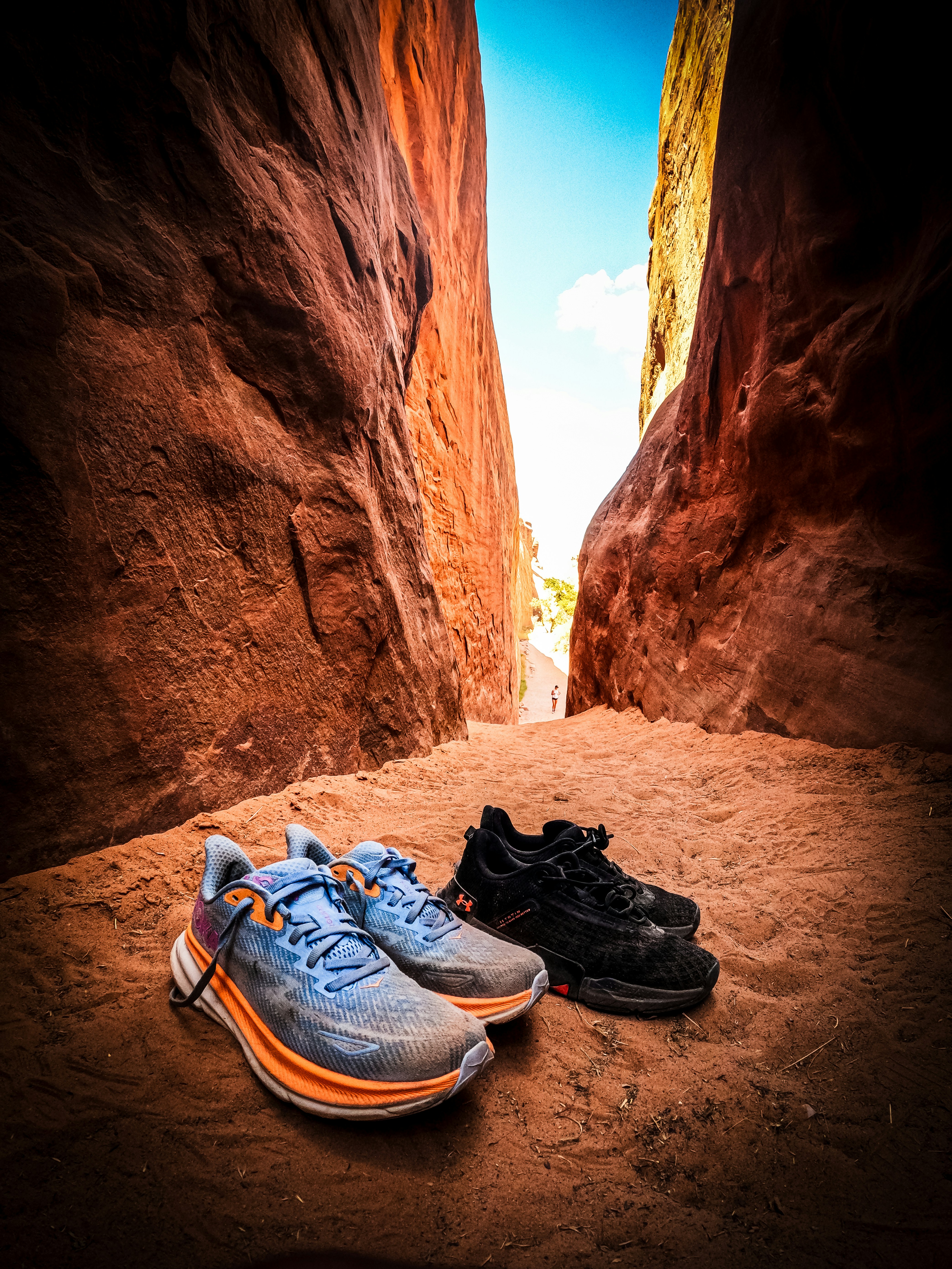 Two sneakers rest on sandy ground between towering red canyon walls, with a bright sky visible through the narrow opening.