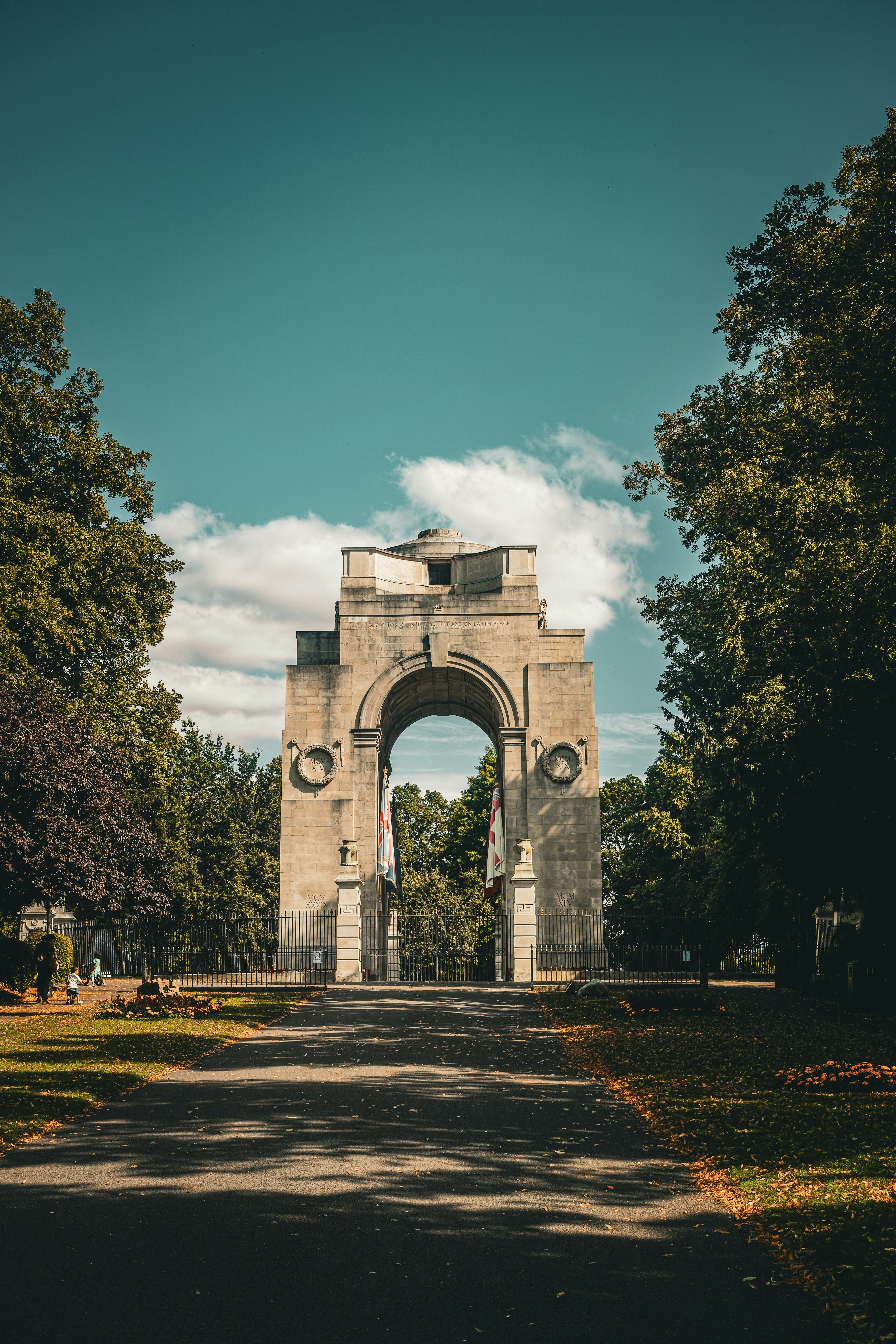 Stone arch gate in a sunlit park, framed by tall trees with a central path leading to the gateway.
