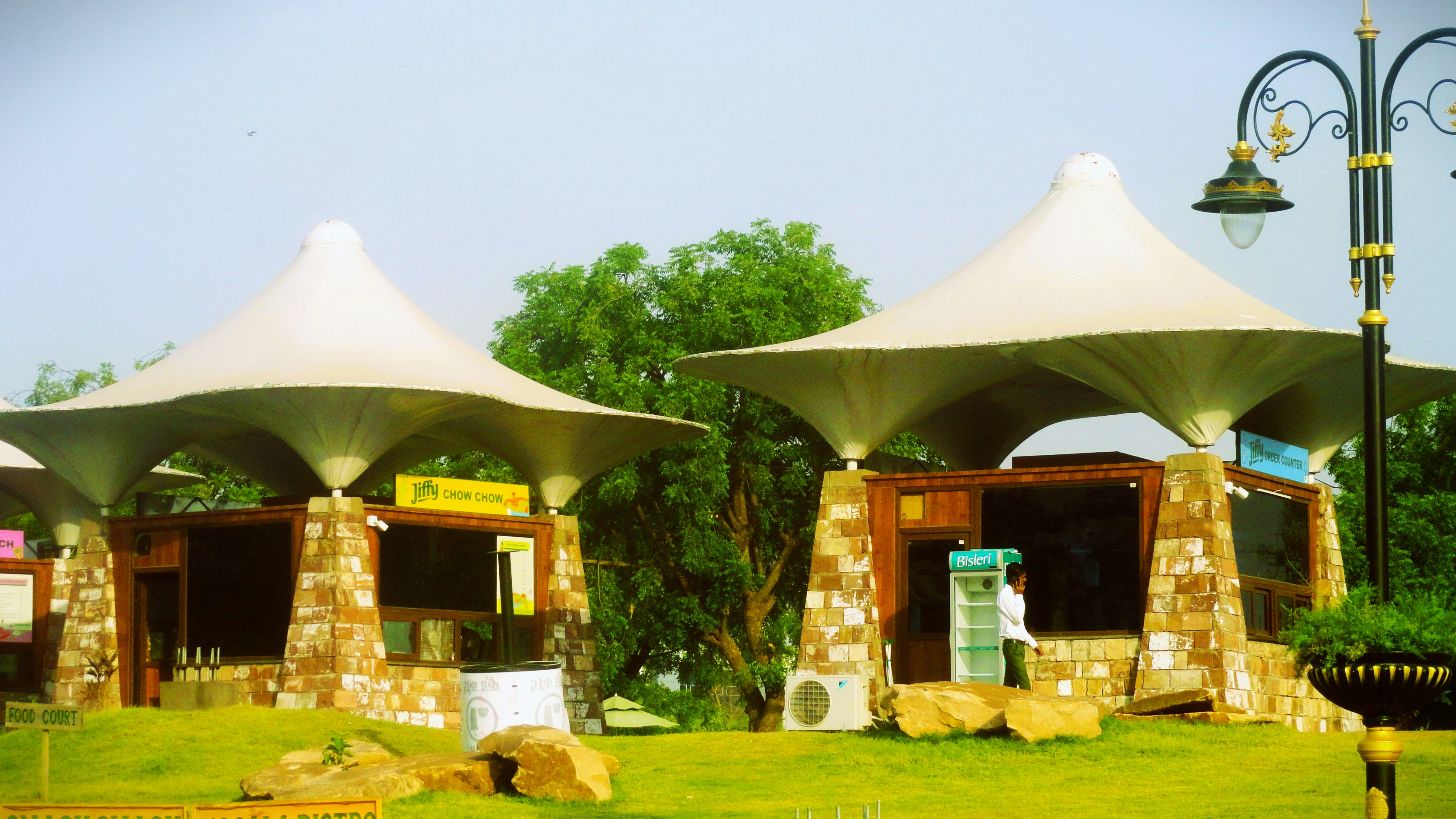 Two brick kiosks rise under white canopy tents in a sunlit park. A person stands near the right pavilion, with a manicured lawn and a decorative lamp visible nearby.