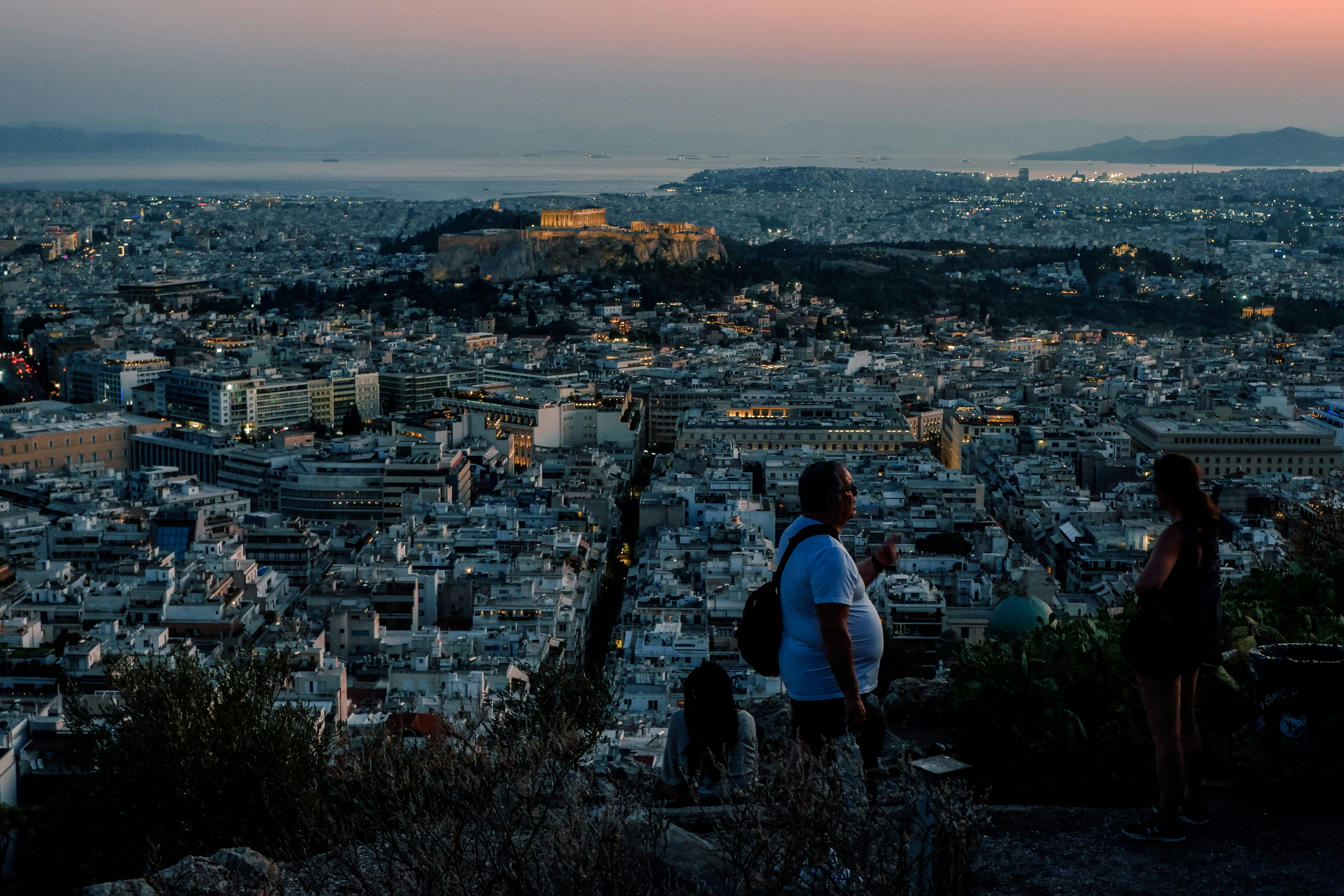 Athens cityscape with the illuminated Acropolis at dusk and silhouettes of people in the foreground.
