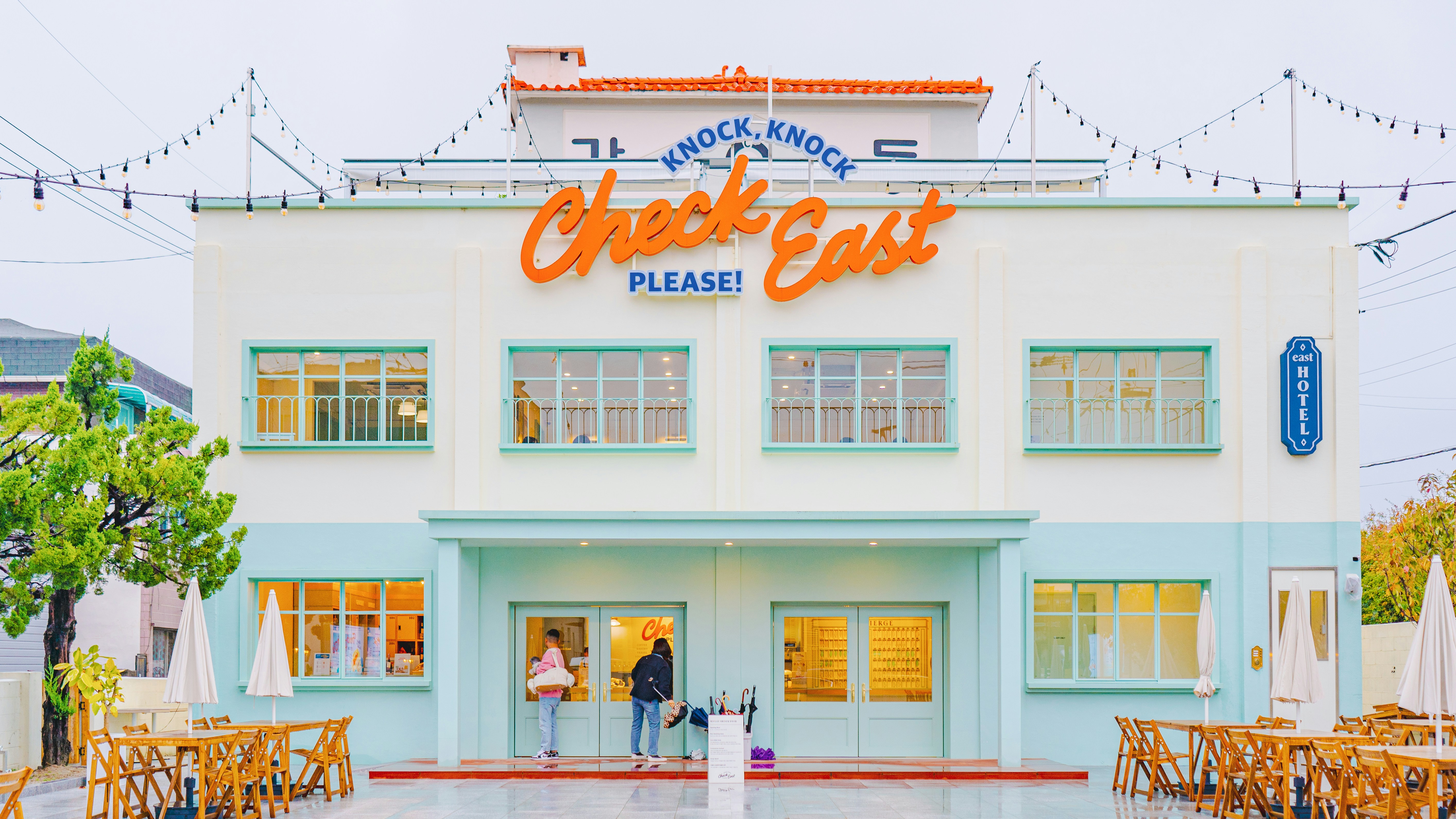 Photograph of a pastel turquoise cafe facade with an orange neon sign reading 'Check East' and string lights overhead. Two people stand at the entrance, inviting customers inside.