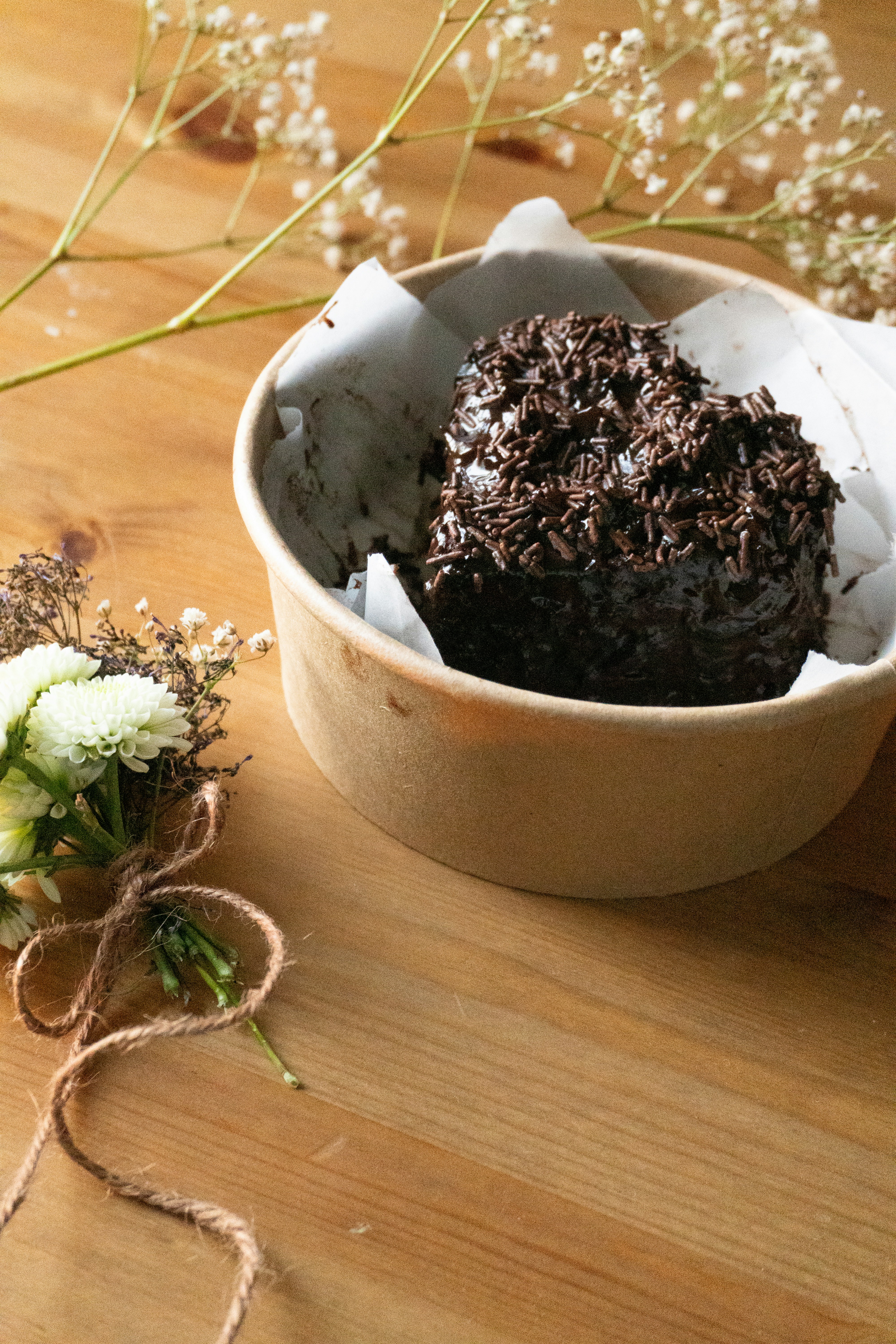 Dark chocolate cupcake in parchment cup, framed by a twine-tied bouquet on a sunlit wooden table.