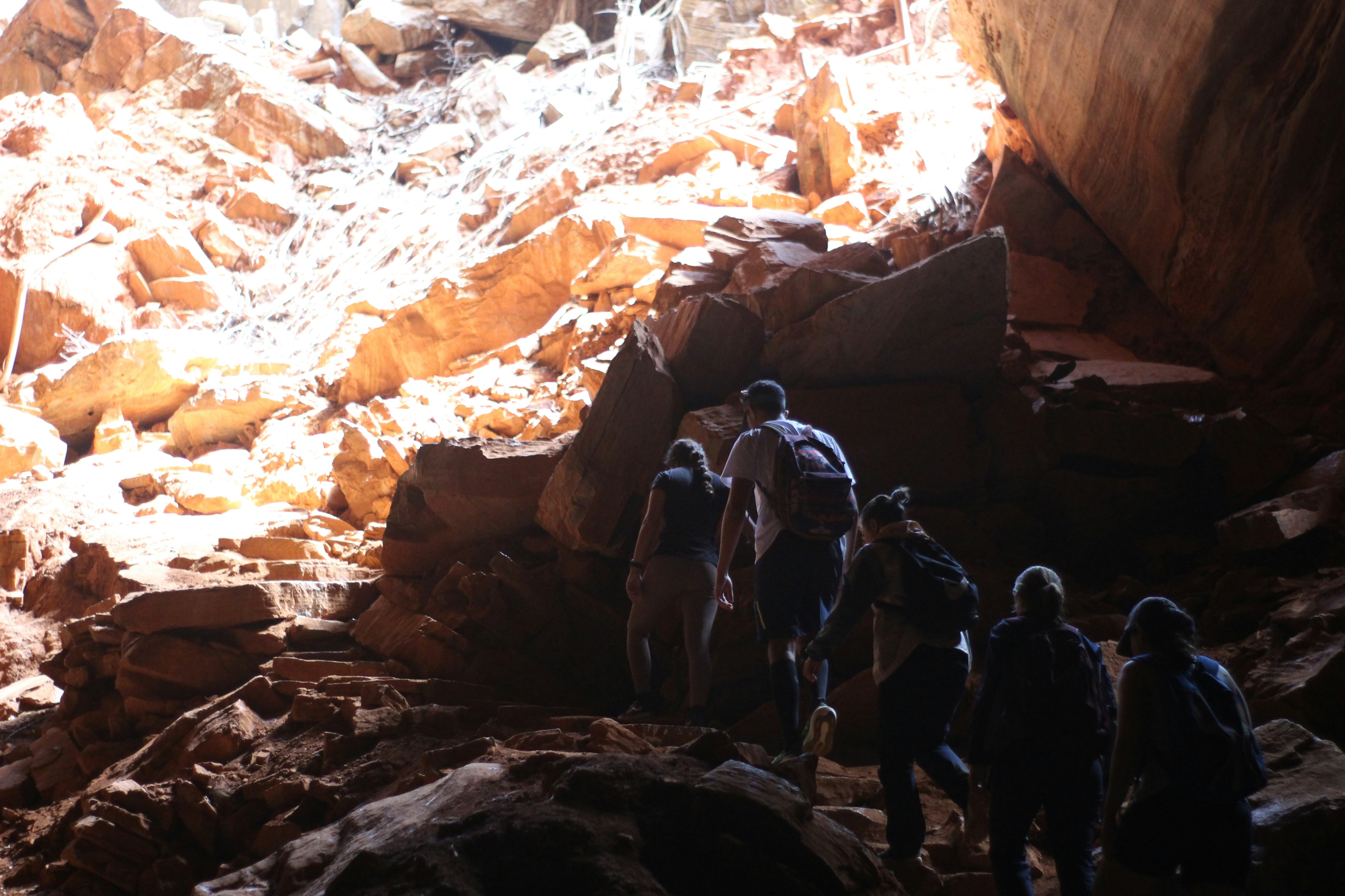 Hikers climb rugged terrain inside a sunlit cave with contrasting shadows.