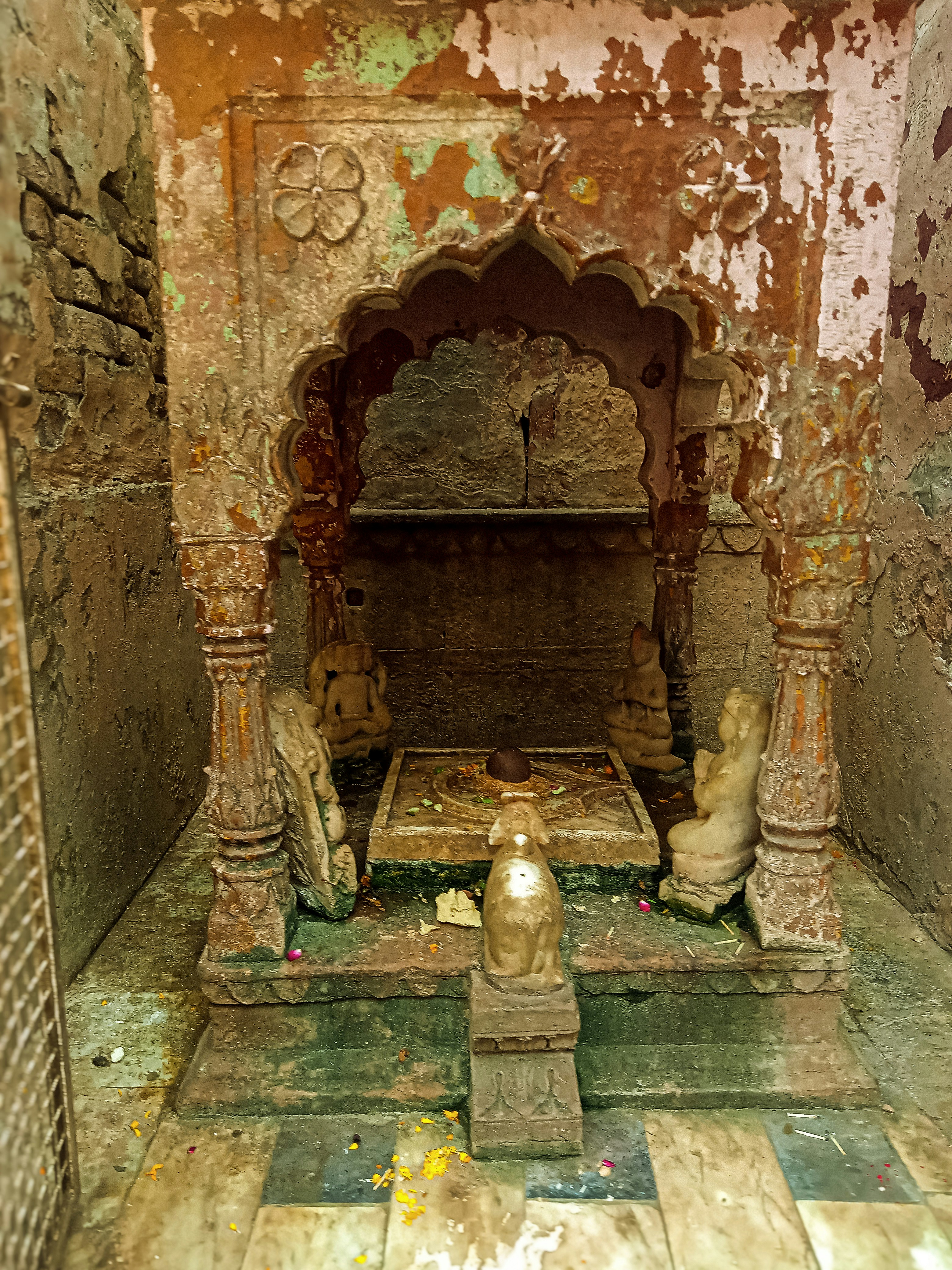 Weathered temple alcove with a sculpted arch, a small central altar, and seated stone figures, set on a tiled floor with scattered petals.