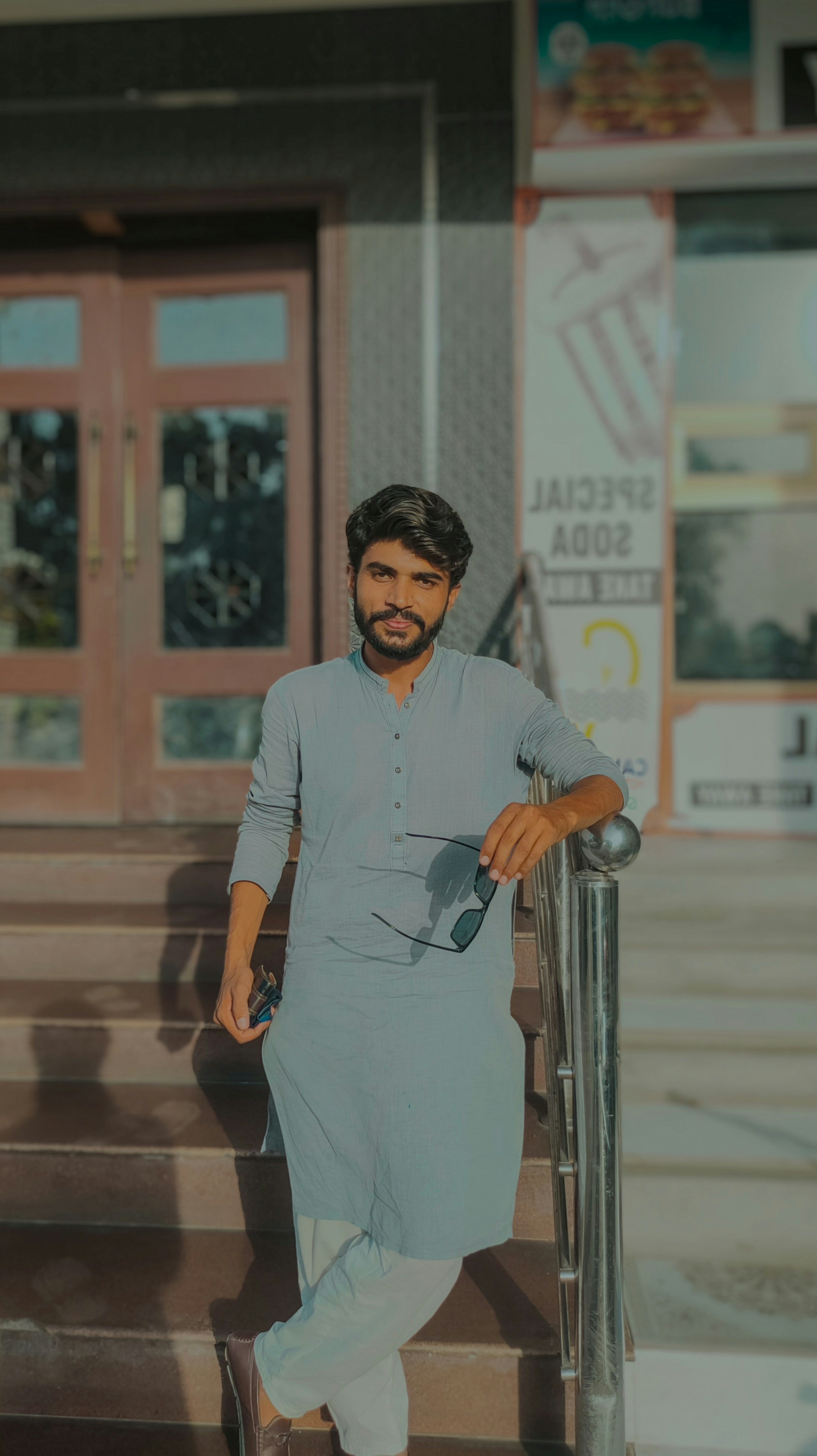 A young man in traditional attire stands confidently on the steps of a building, holding sunglasses. The warm sunlight casts soft shadows, enhancing the scene's tranquility.