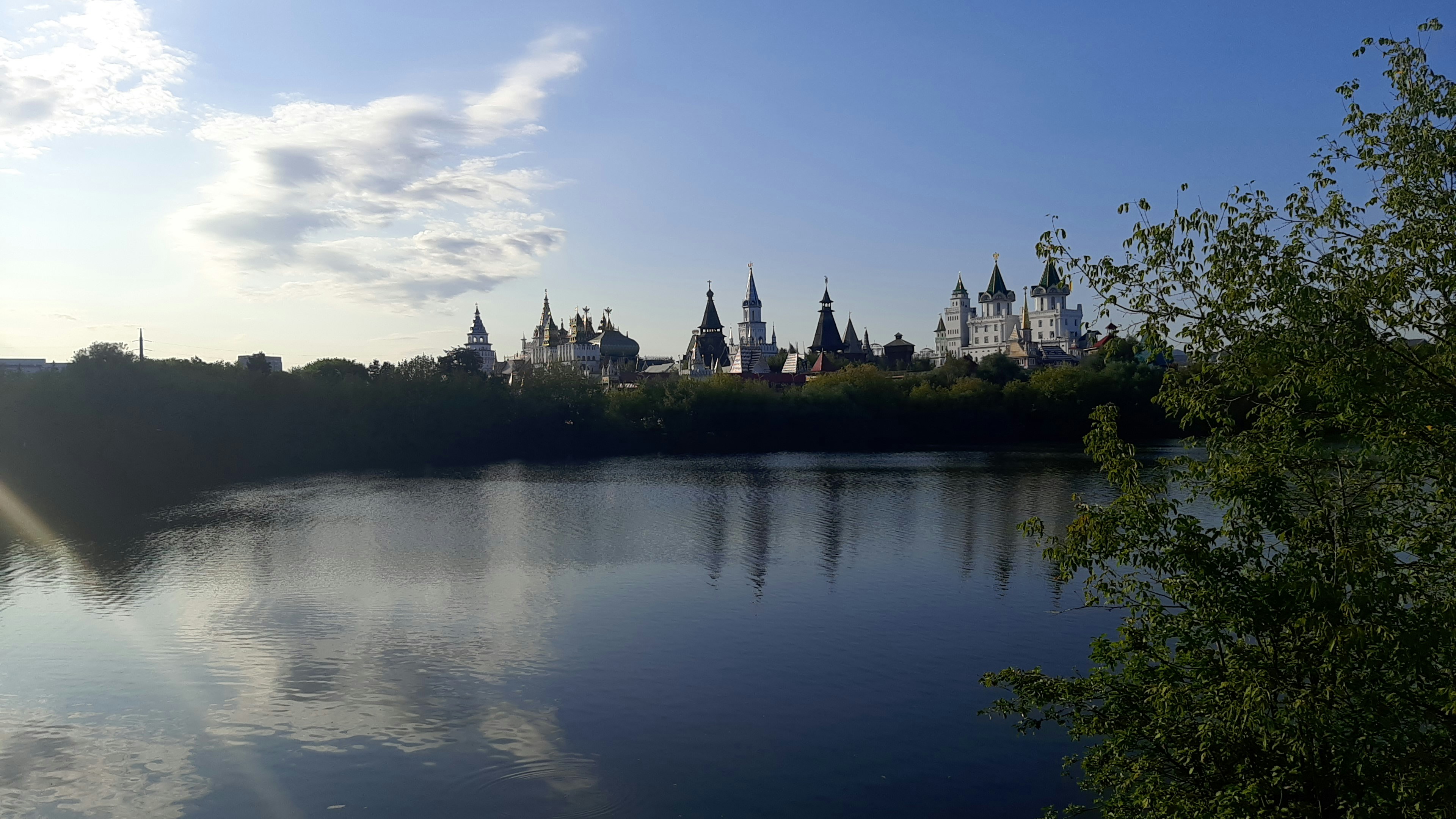 Dawn over a calm river mirrors a skyline of spired buildings along the horizon. Trees frame the foreground on the right.