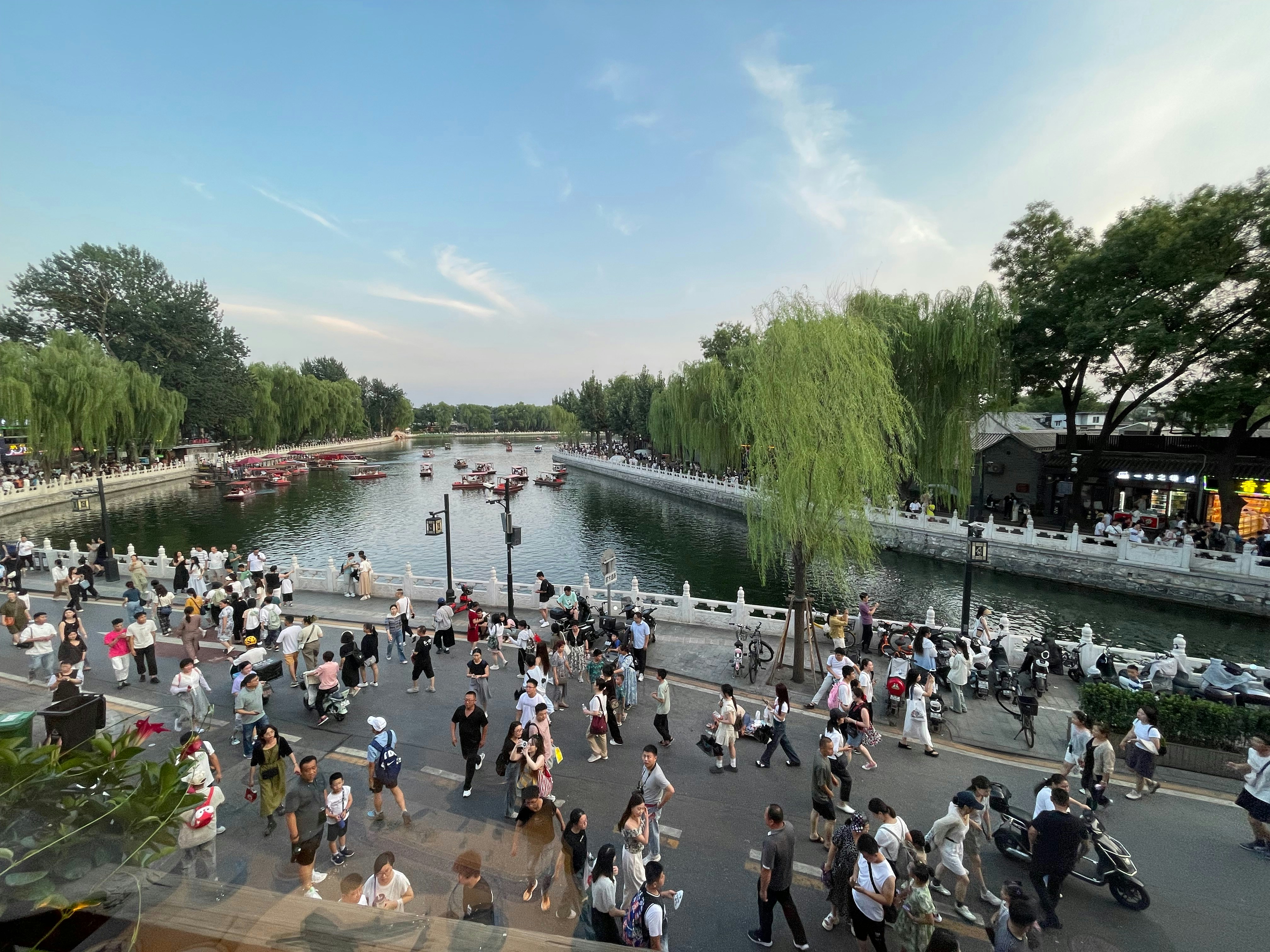 Bustling riverside scene filled with people enjoying leisurely activities, surrounded by lush greenery and boats on the water.
