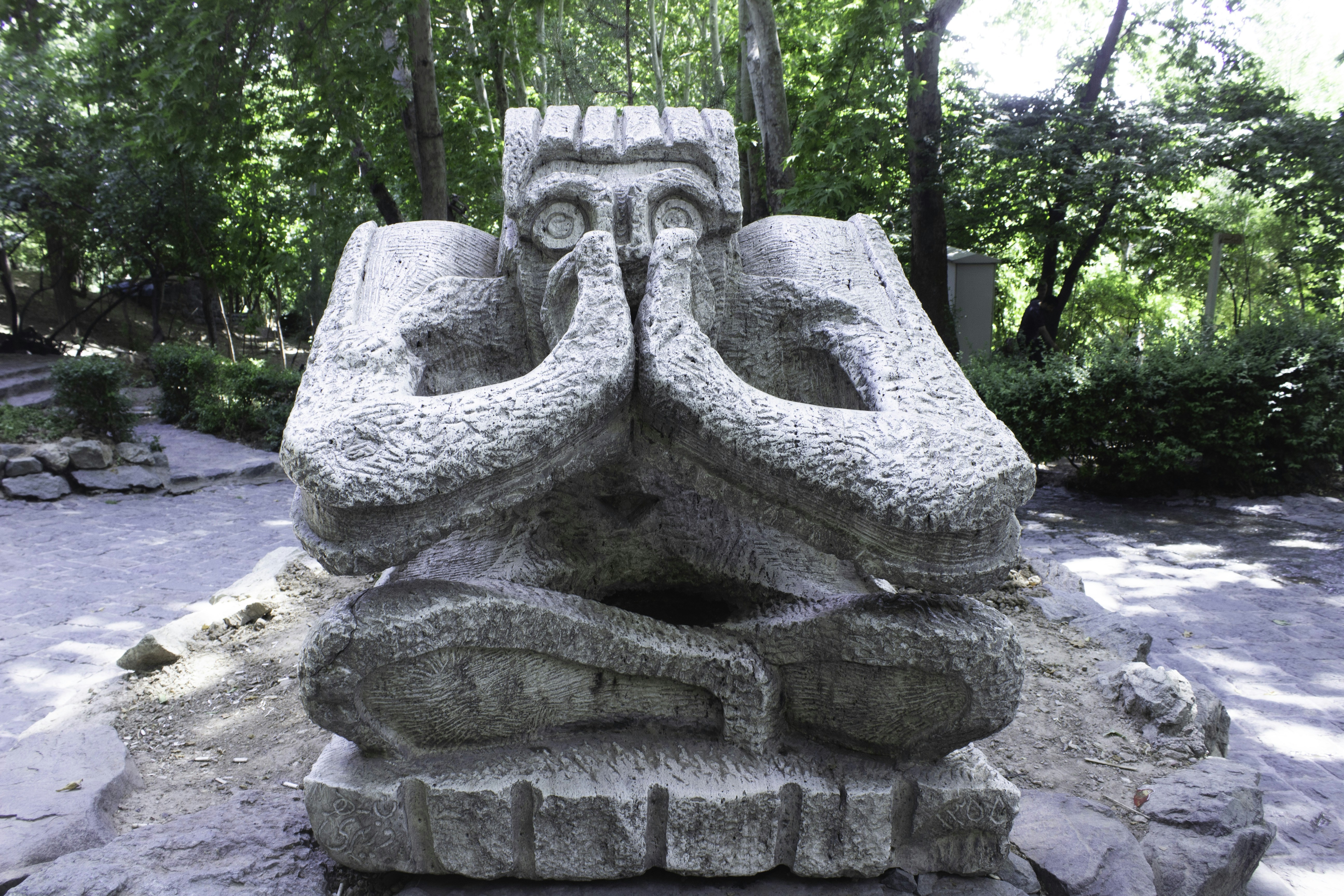 Intricate stone sculpture depicting a seated figure in a meditative pose, surrounded by lush greenery. The artwork invites contemplation.