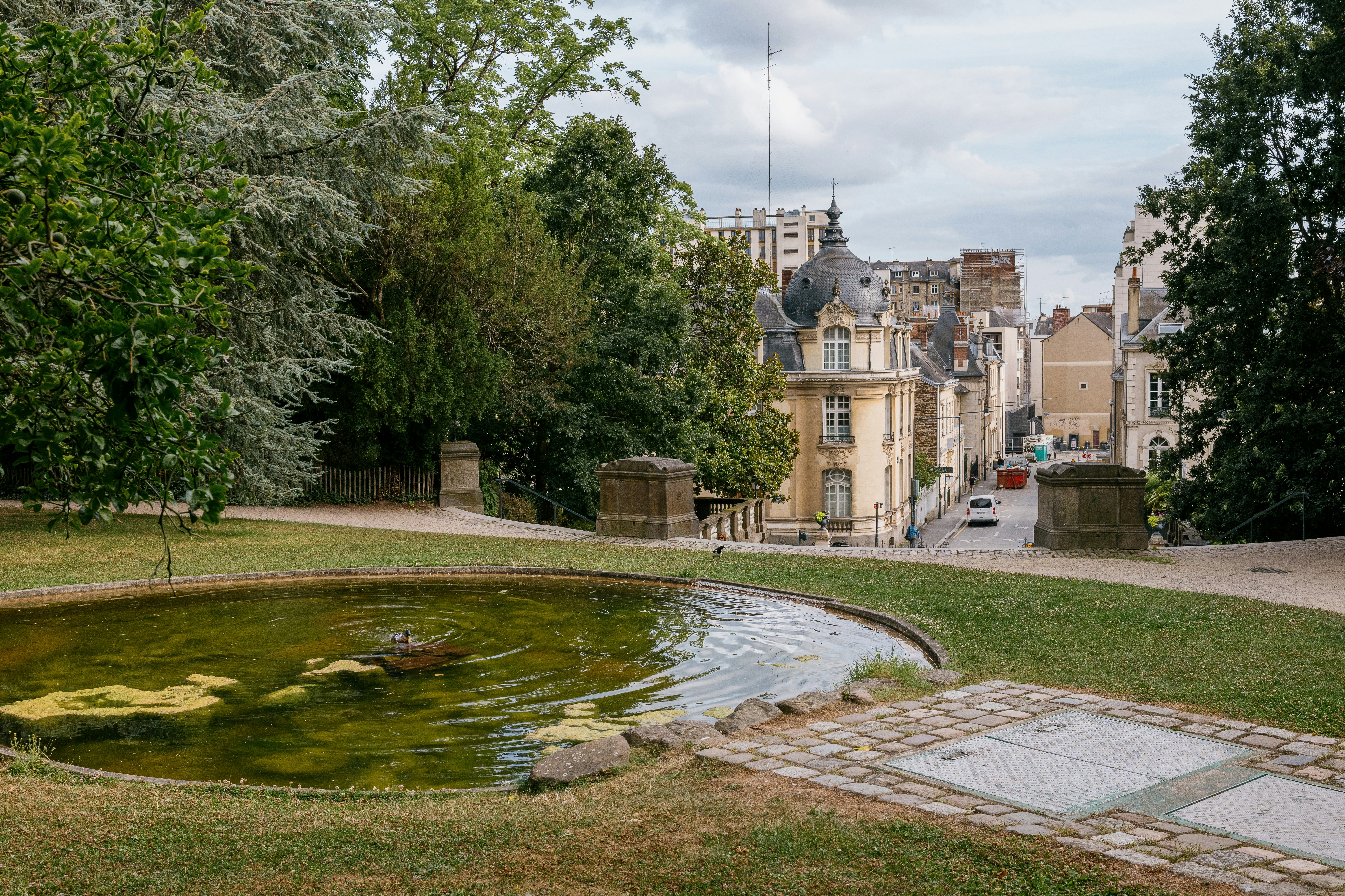 Small pond with surrounding greenery and distant view of historic Rennes architecture under a cloudy sky.