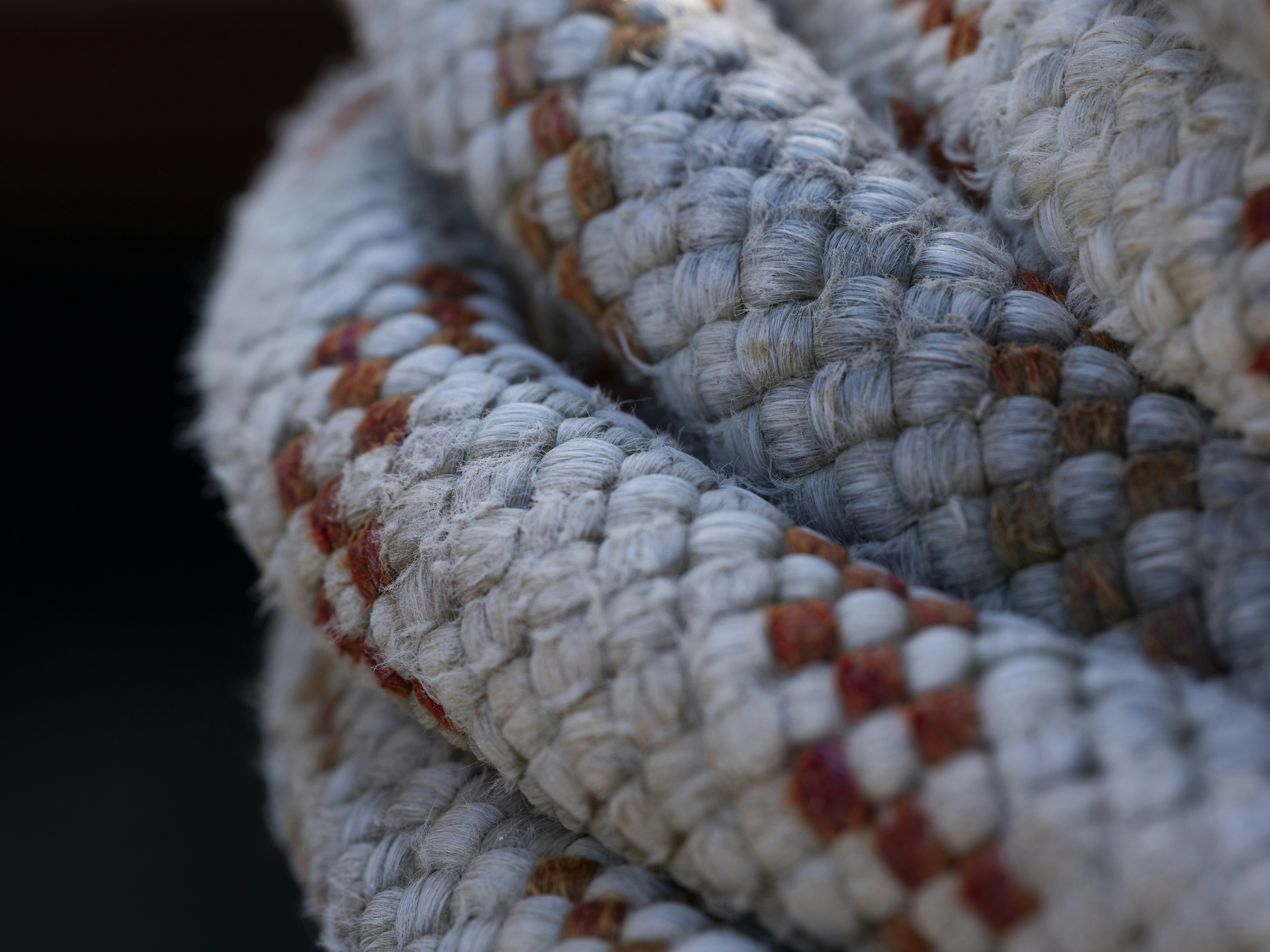Close-up of braided rope with light gray fibers and red accents, showing tight weave and gentle coil forms. The shallow depth of field isolates texture against a dark background.