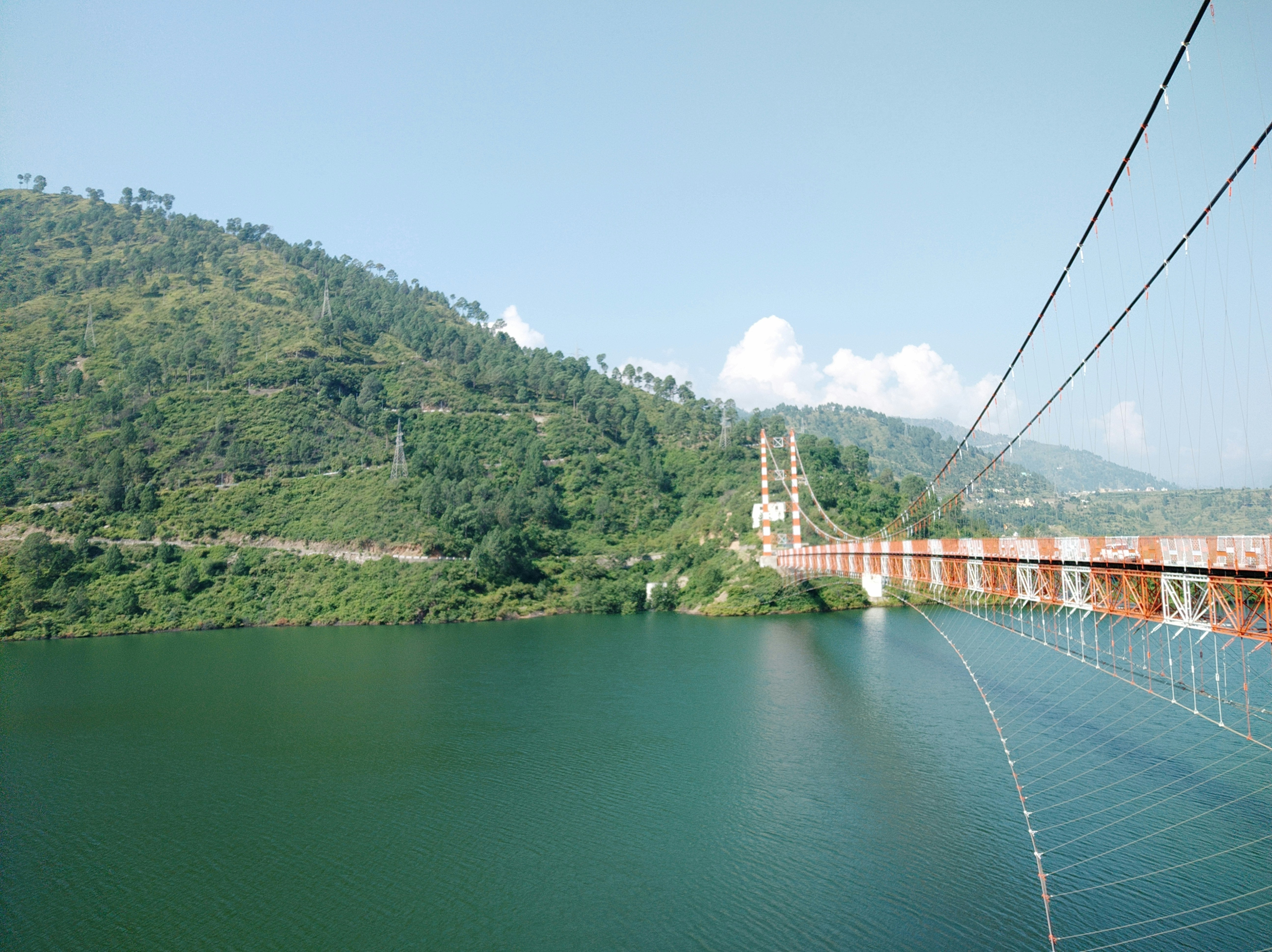 Landscape photograph of a suspension bridge spanning an emerald-green lake with forested hills in the background.
