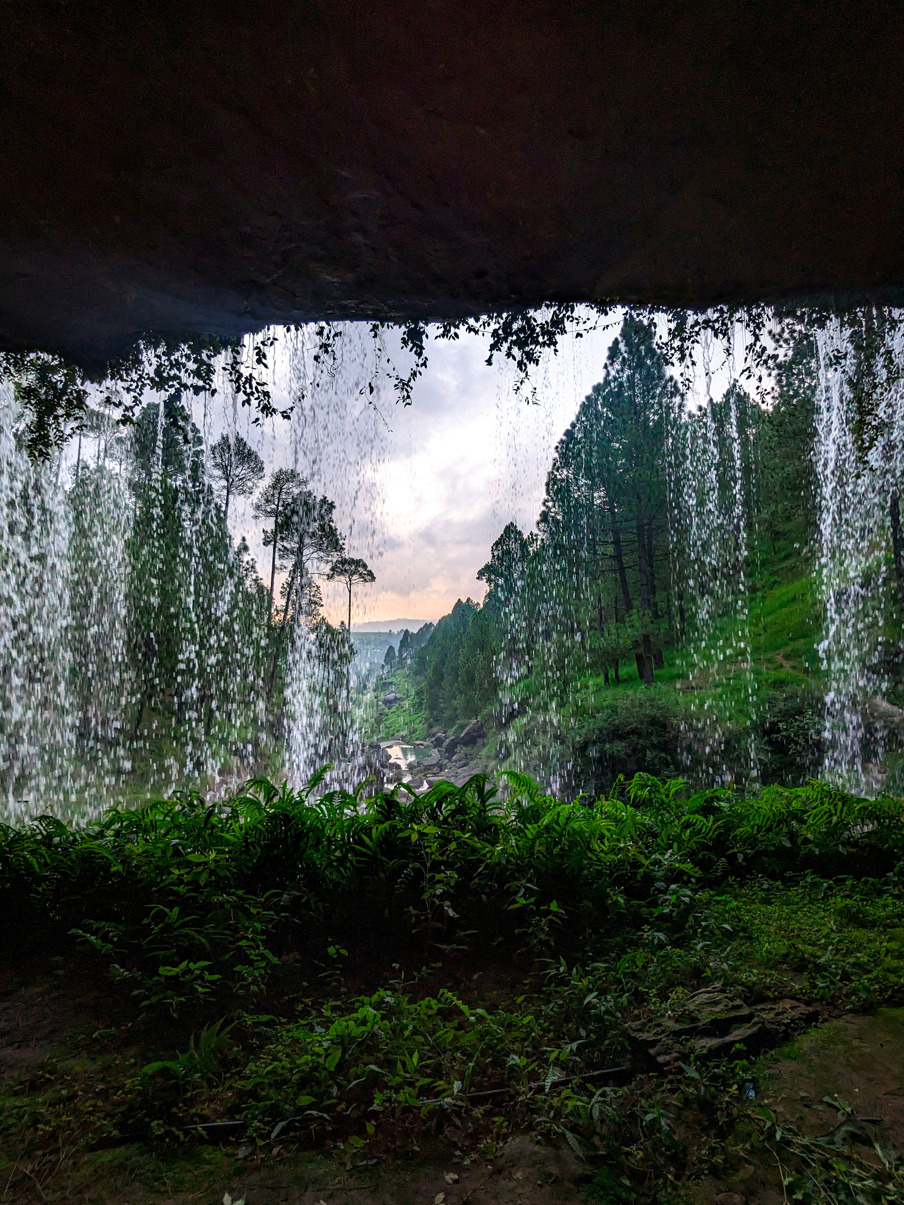 Dark cave opening with water curtains streaming down from above. Beyond the veil, a lush green valley and distant river unfold in soft light.