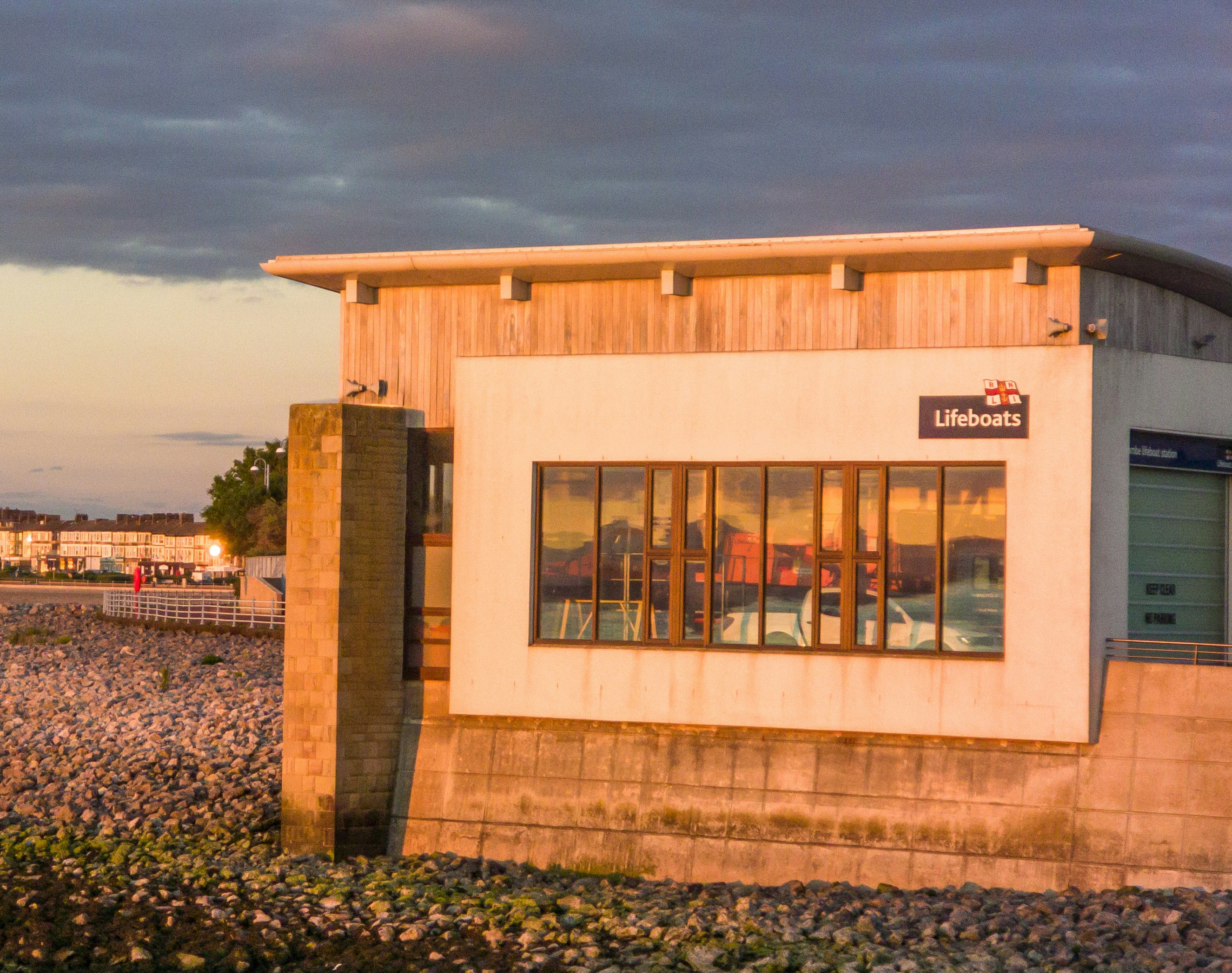 Lifeboat station with a broad window facade along a pebble beach. The glass reflects warm hues, emphasizing maritime architecture.