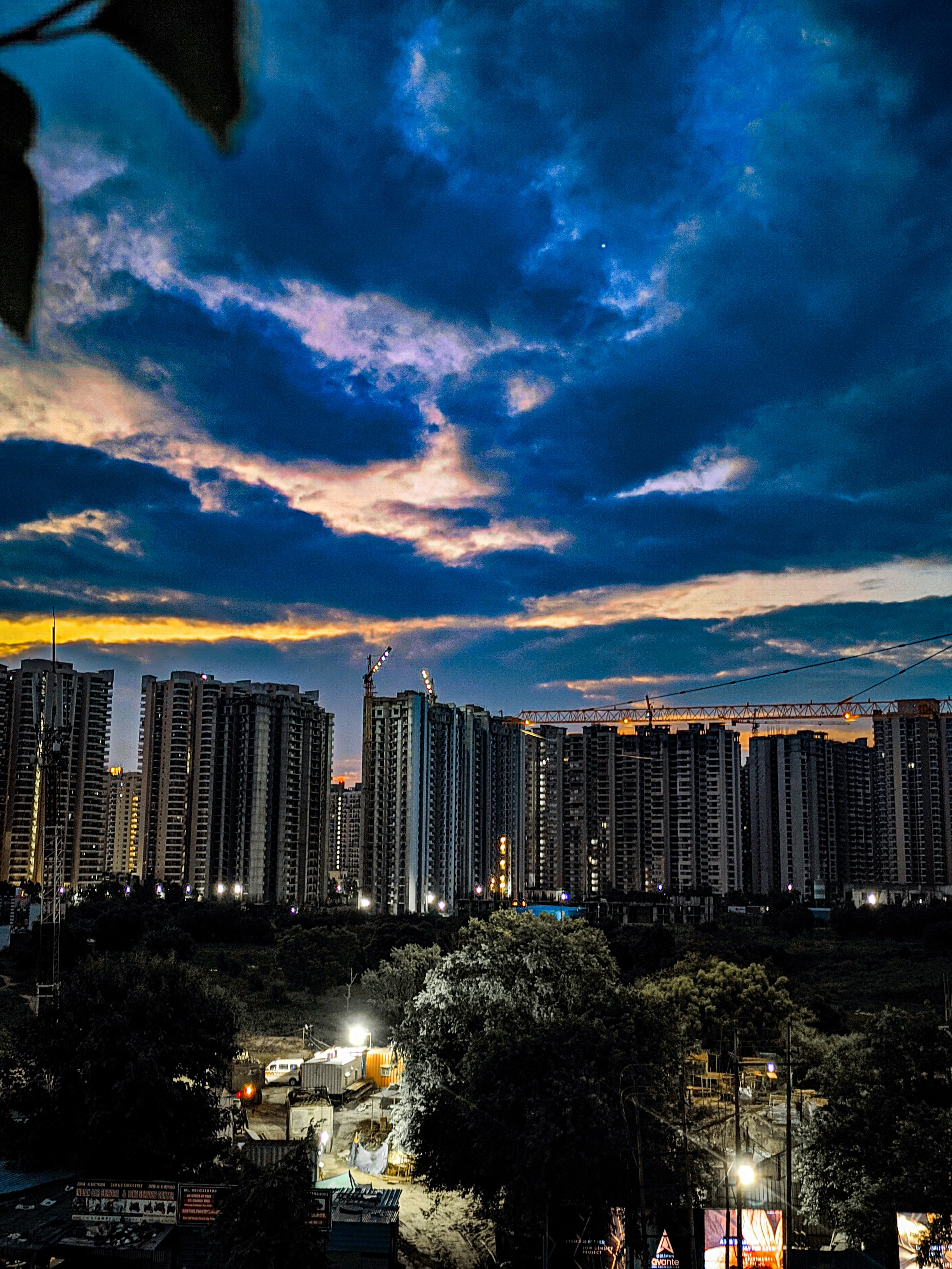 City skyline illuminated against a dramatic twilight sky, with clouds reflecting hues of orange and blue. Construction cranes stand tall amidst the urban landscape.
