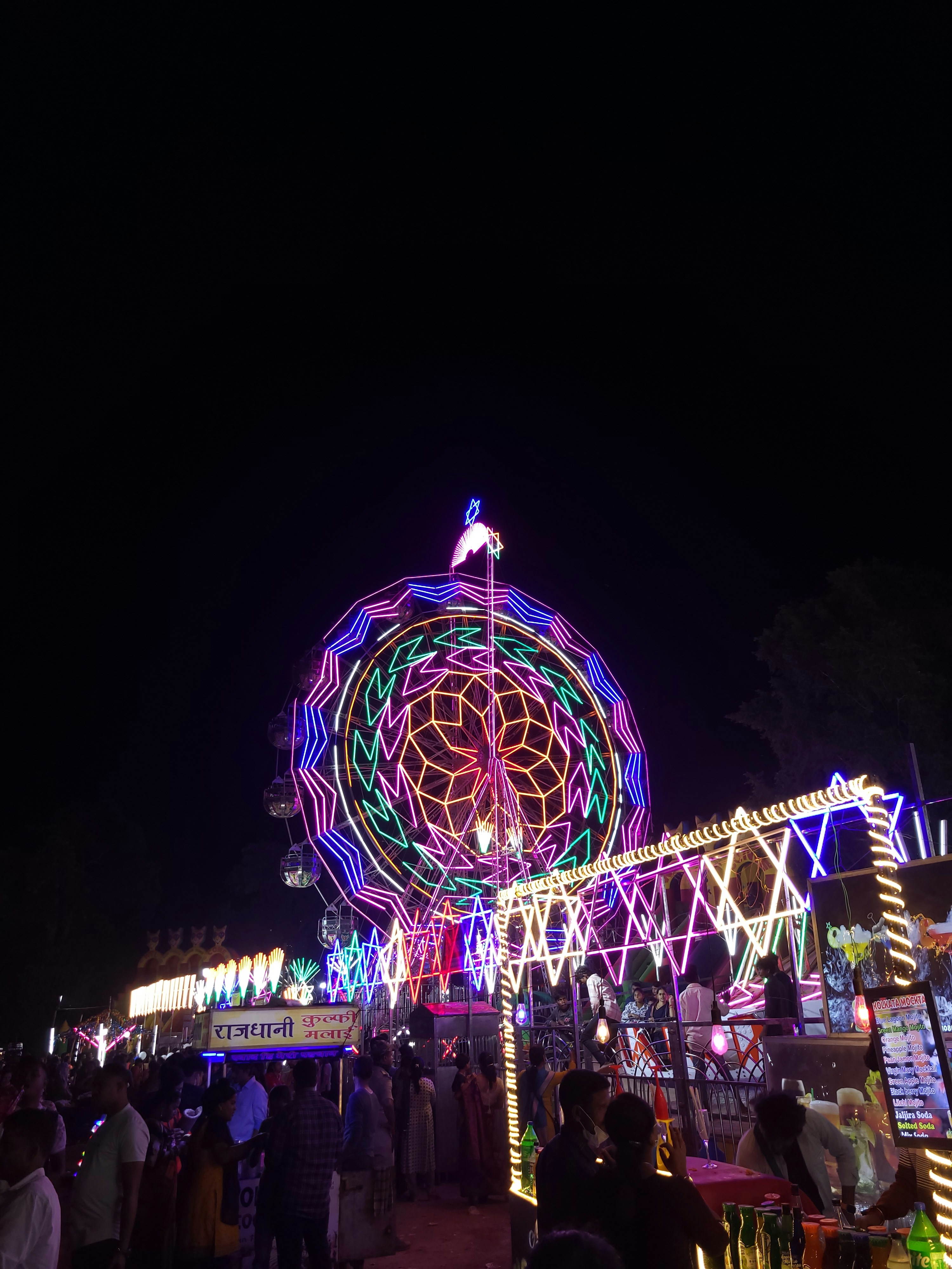 Nighttime carnival scene with a large neon-lit Ferris wheel and geometric rainbow lights, crowds milling around bright stalls.