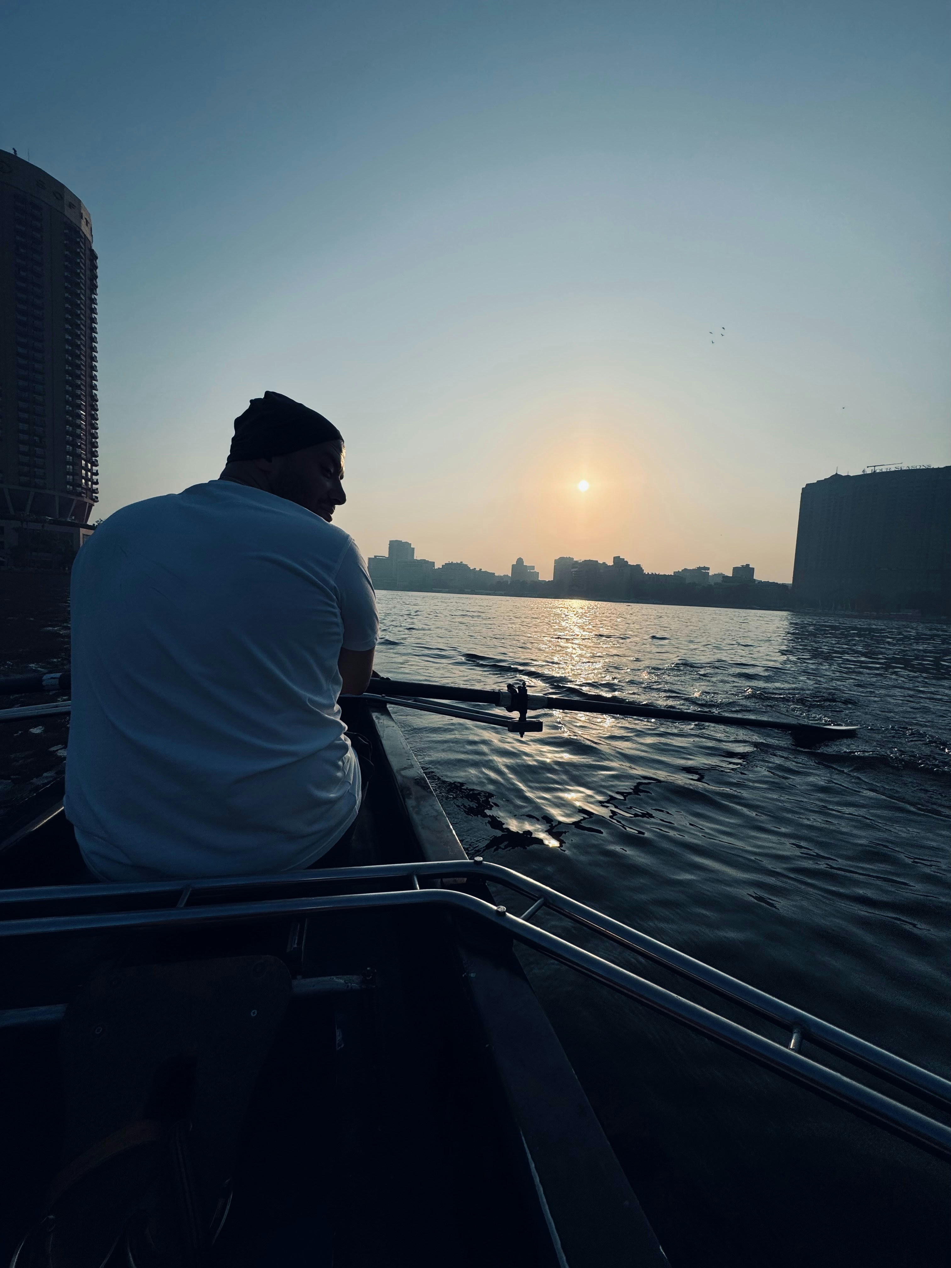 Silhouetted rower gazing over a calm river at sunrise, surrounded by city buildings.