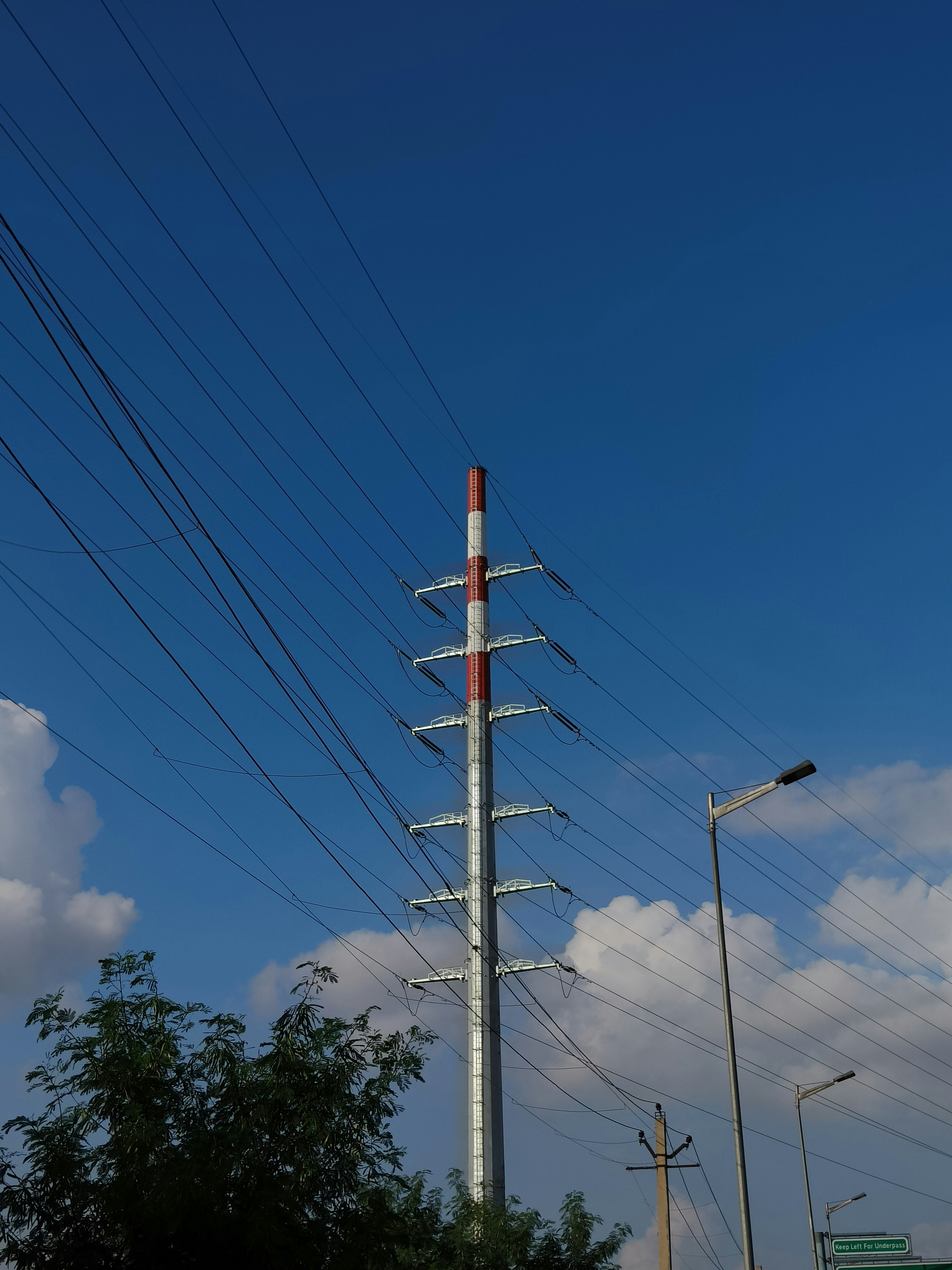 A tall concrete utility pole with red-and-white markings rises among a tangle of power lines. Street lamps line the right edge against a crisp blue sky.