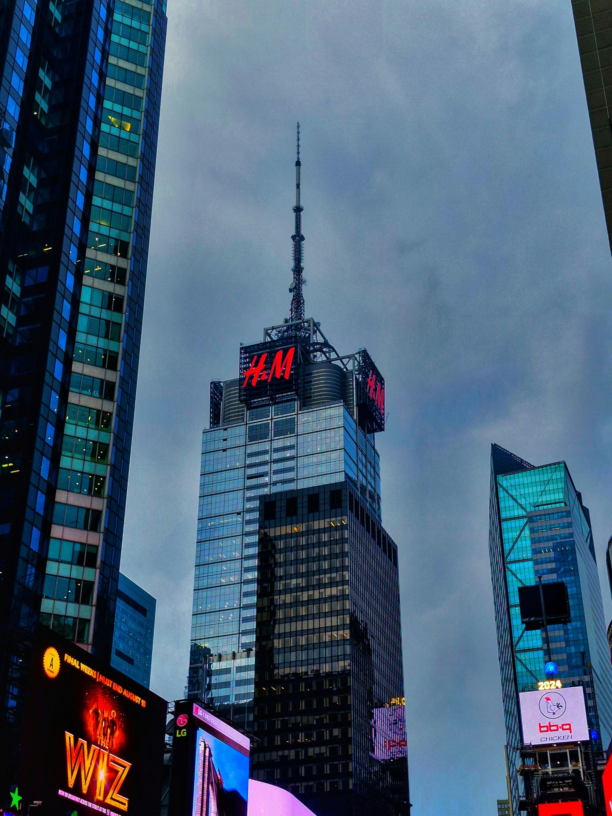 City skyline at dusk with glass towers and neon billboards. The central building features a prominent red H&M logo and antenna.