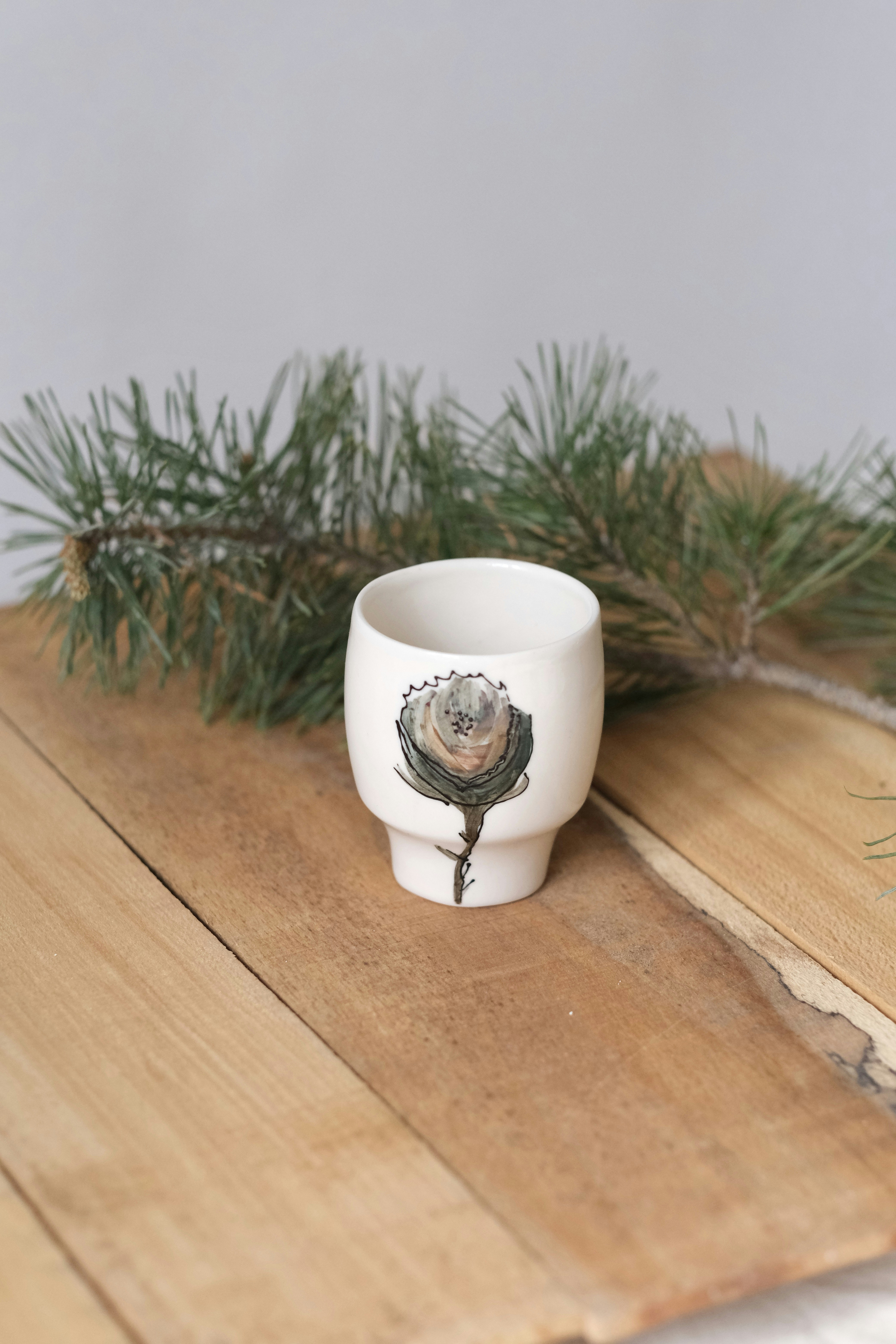 Ceramic cup adorned with a watercolor flower rests on weathered wooden boards, with pine sprigs providing a natural backdrop.