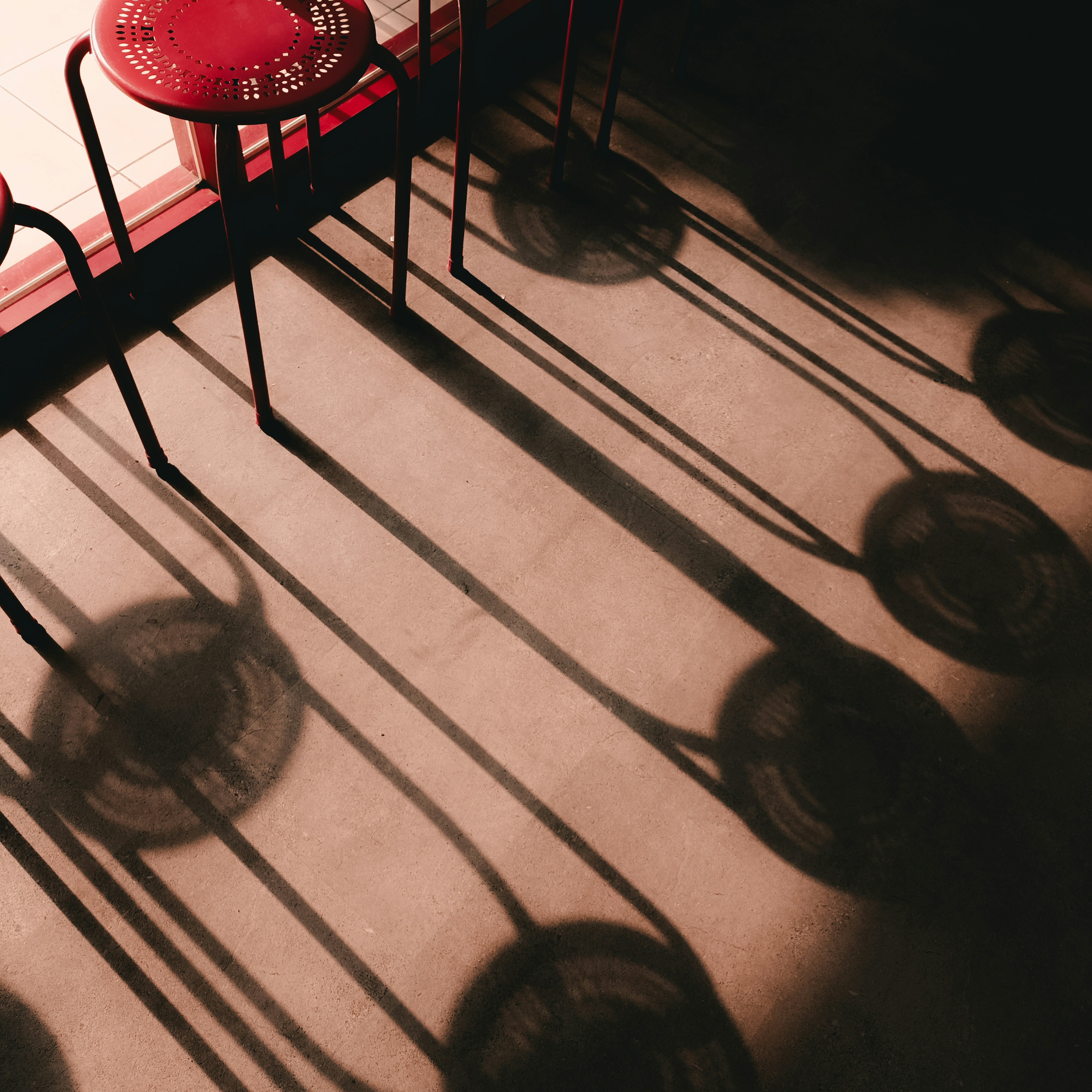 A red perforated chair sits near a window, its long shadows stretching across a concrete floor, creating stark geometric patterns.