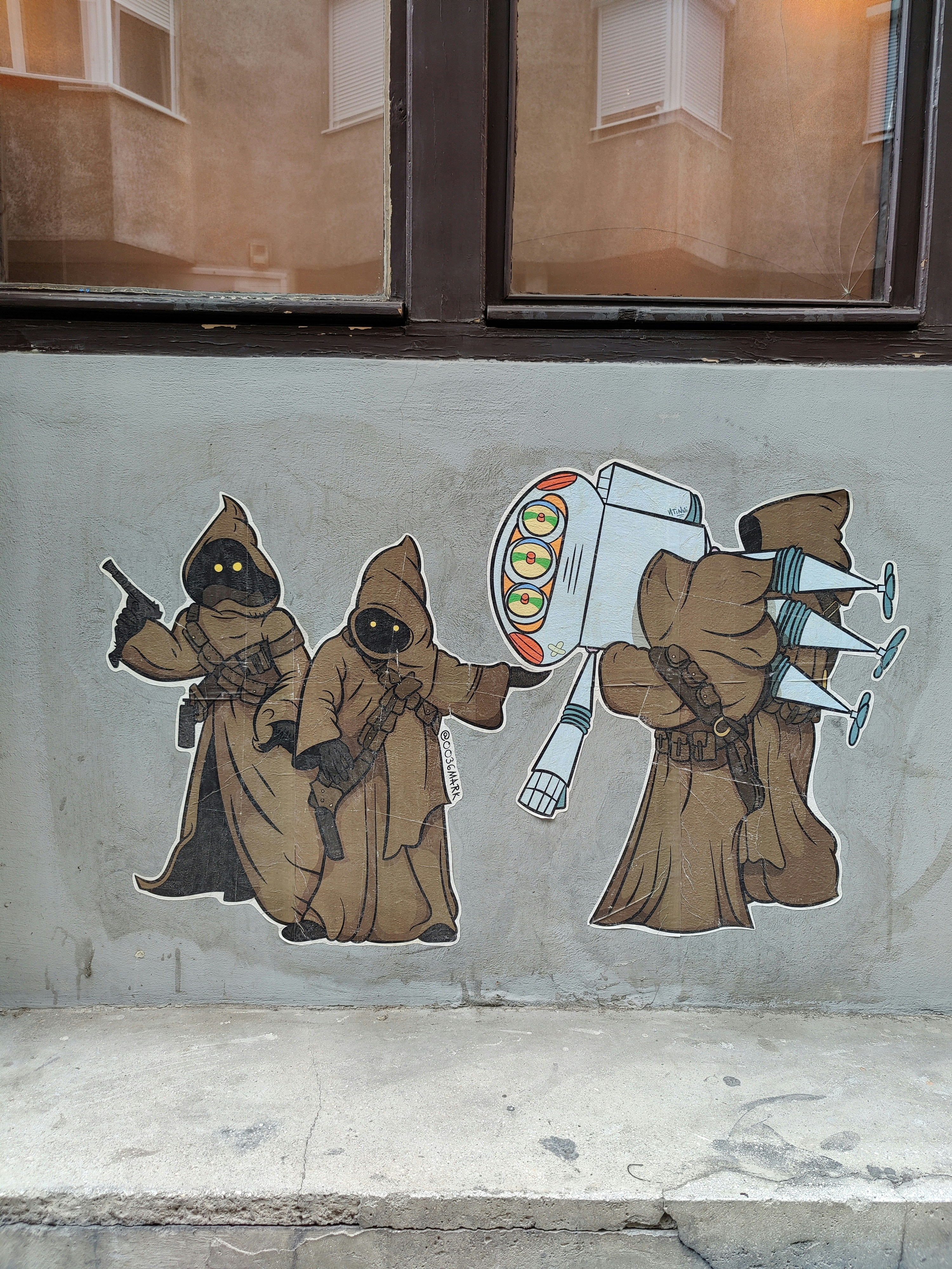 Mural painting of three hooded figures in brown robes on a gray wall, with one holding a colorful circular orb and another carrying a large mechanical gadget.