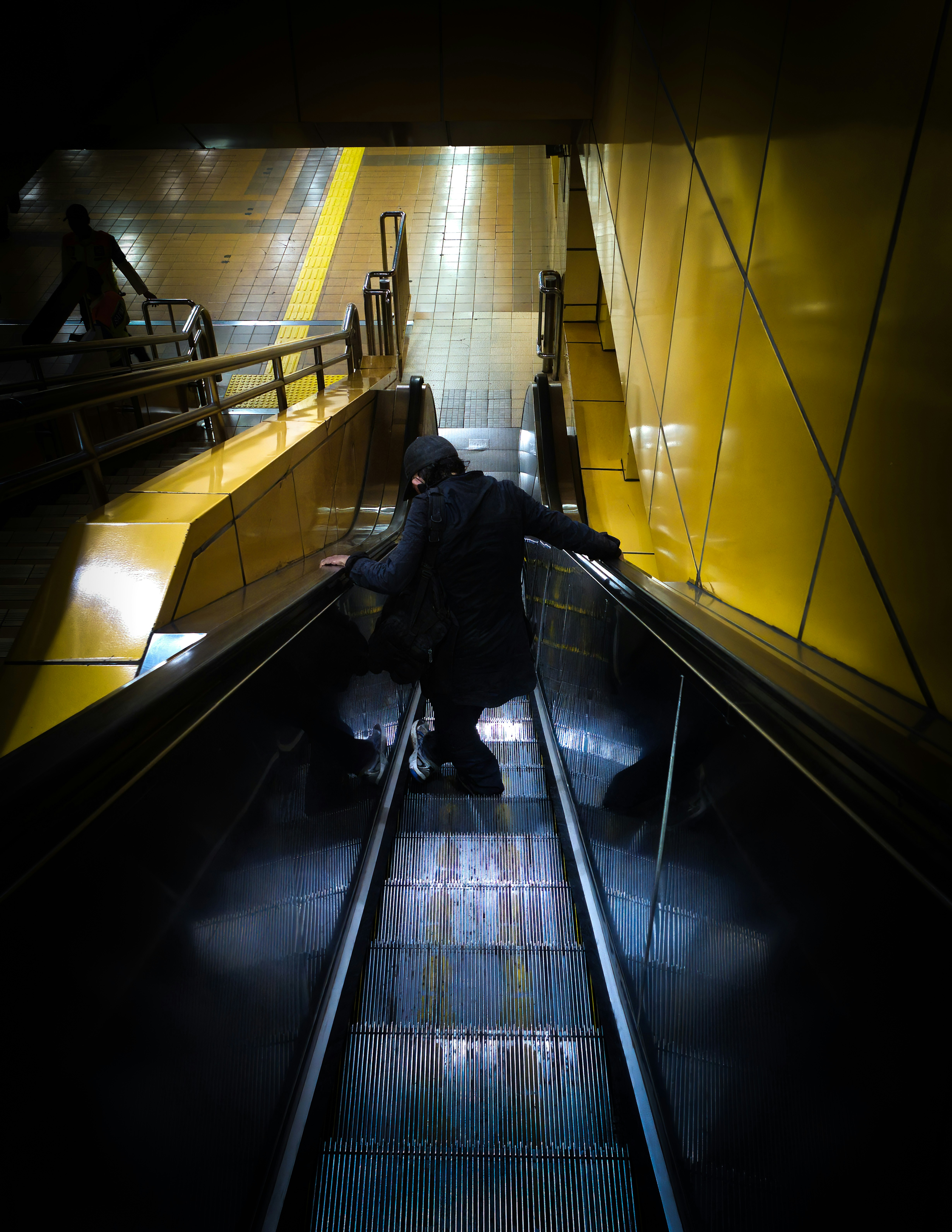 A solitary traveler in dark clothing descends a yellow-walled escalator in a dim, urban transit setting.