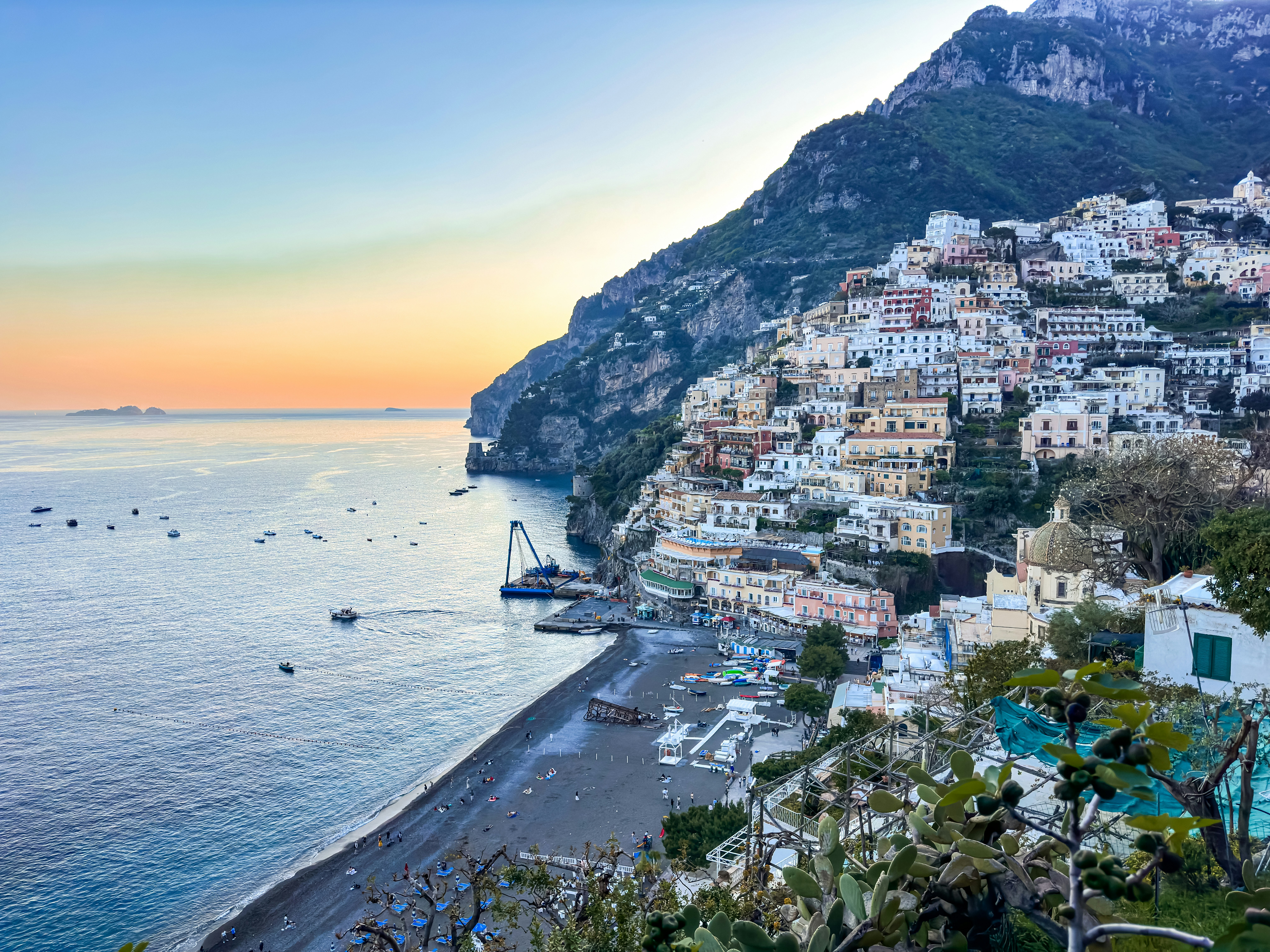 Colorful hillside town overlooking a tranquil sea at sunset, with boats scattered across the water.