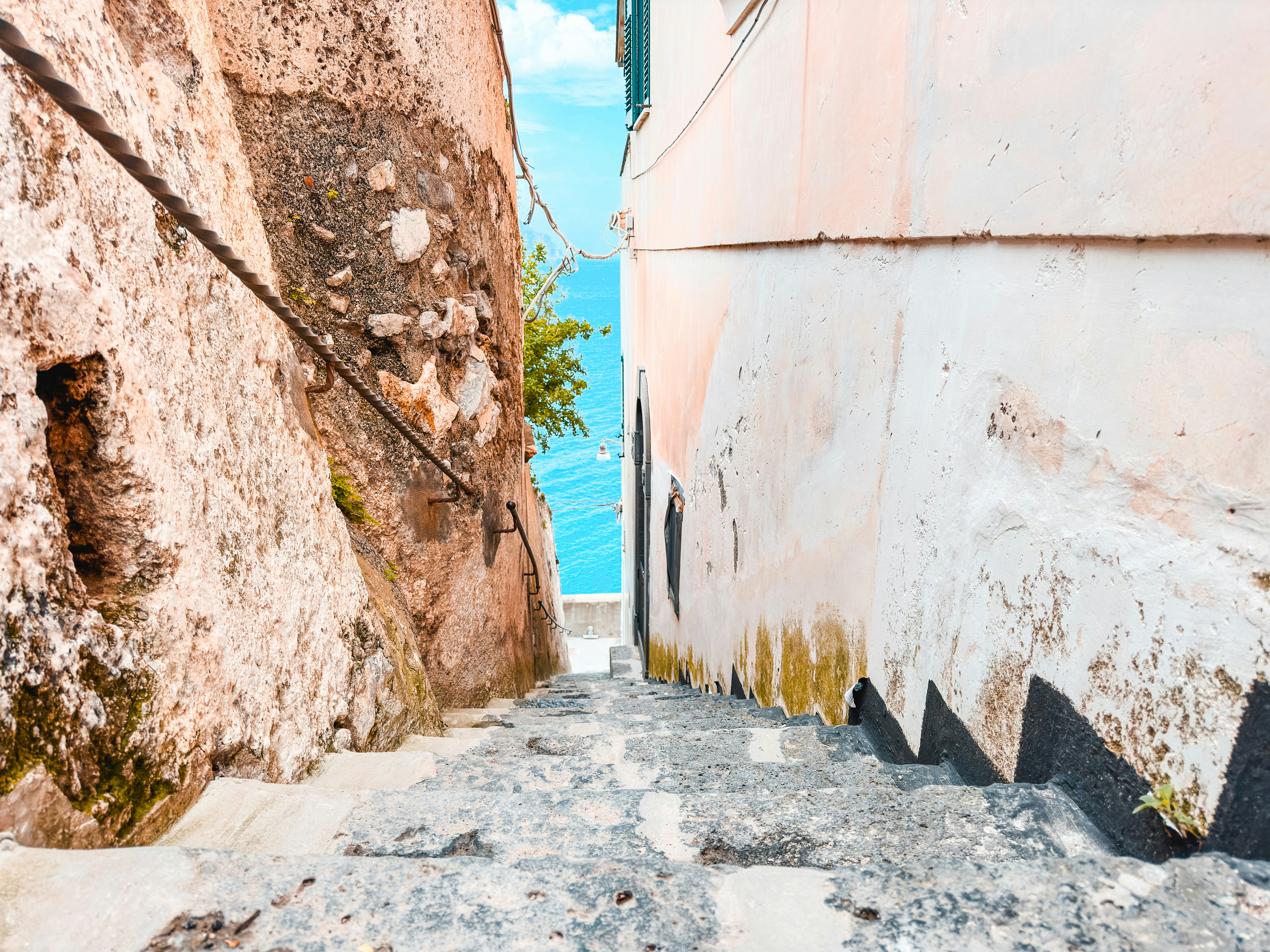 Narrow stone staircase leading down between weathered walls towards a vibrant blue sea under a clear sky.