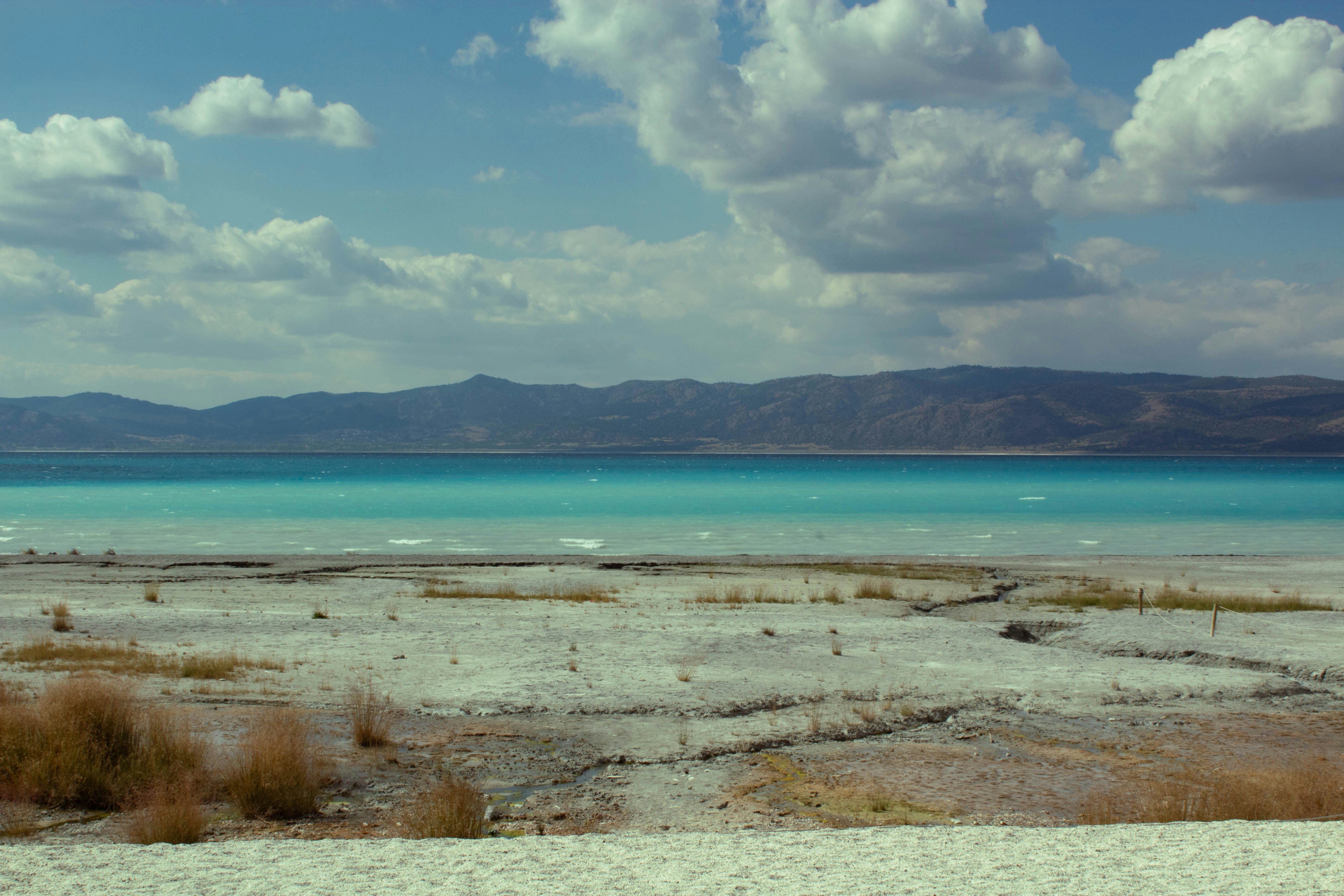 Tranquil turquoise lake bordered by a sandy shore with distant mountains under a cloud-filled sky.
