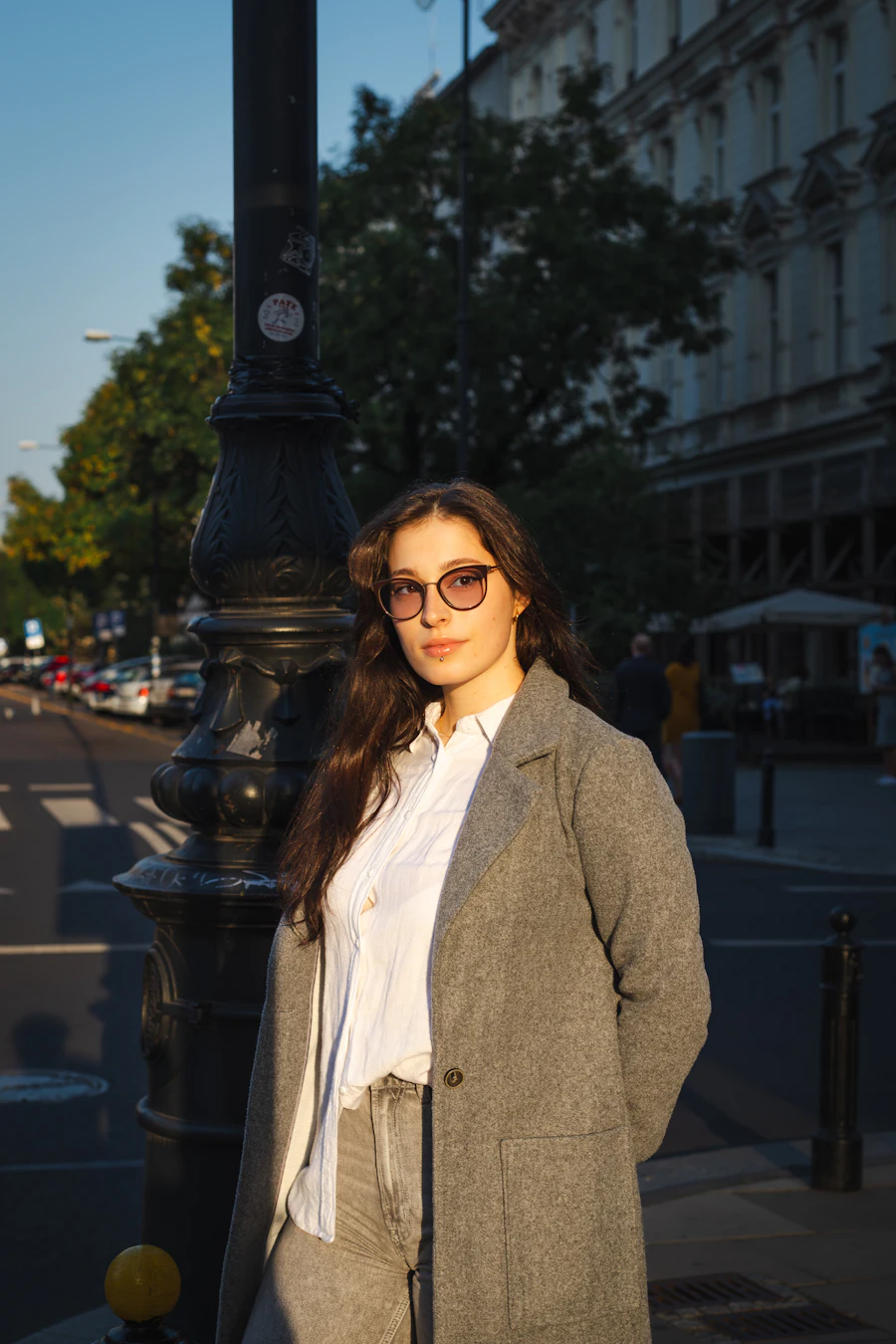 Woman in a gray coat and glasses standing beside a decorative lamppost on a sunlit urban street, with buildings, trees, and traffic in the background.