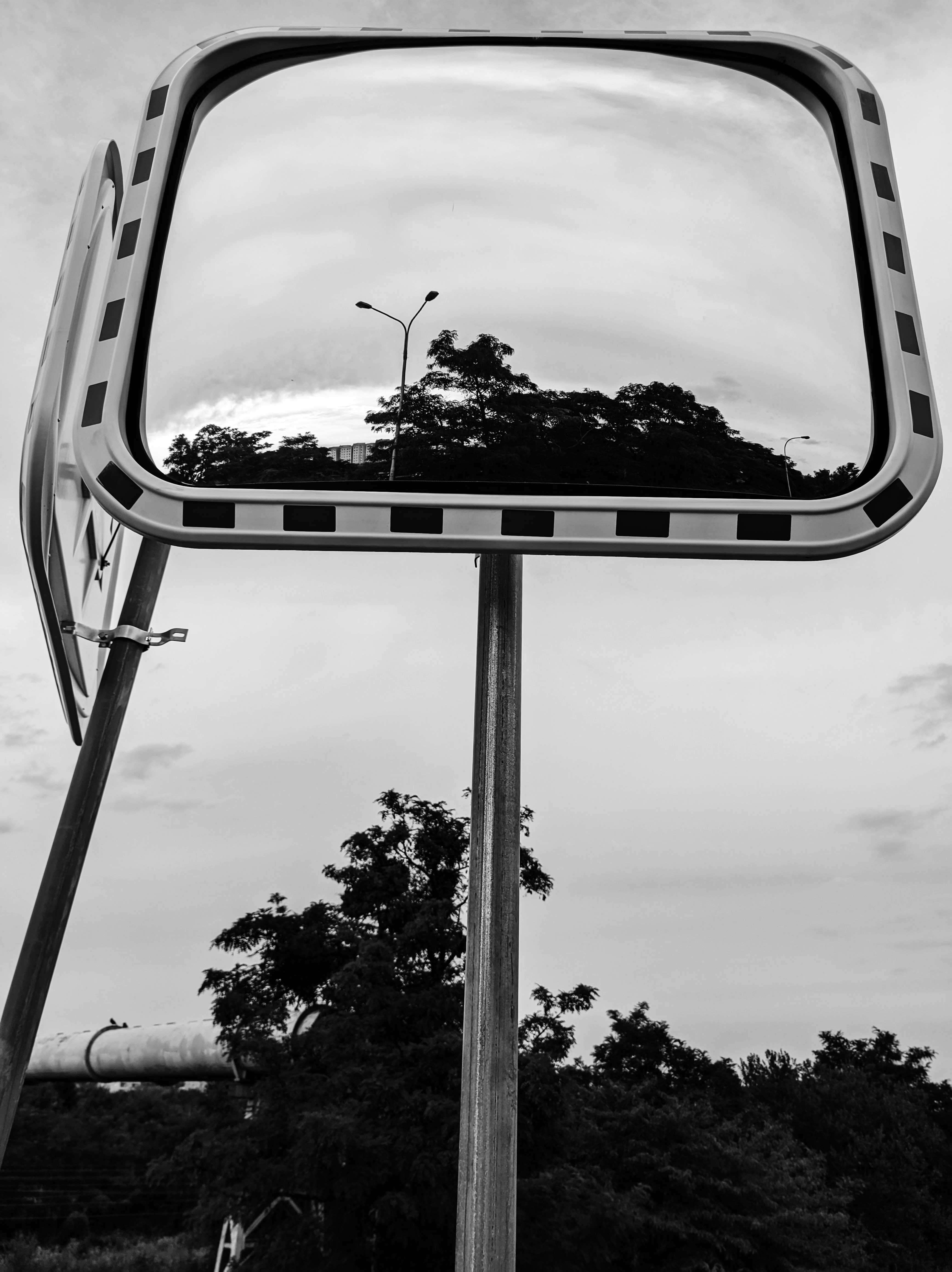 Monochrome photograph of a curved roadside mirror reflecting silhouetted trees and a streetlamp against a cloudy sky.