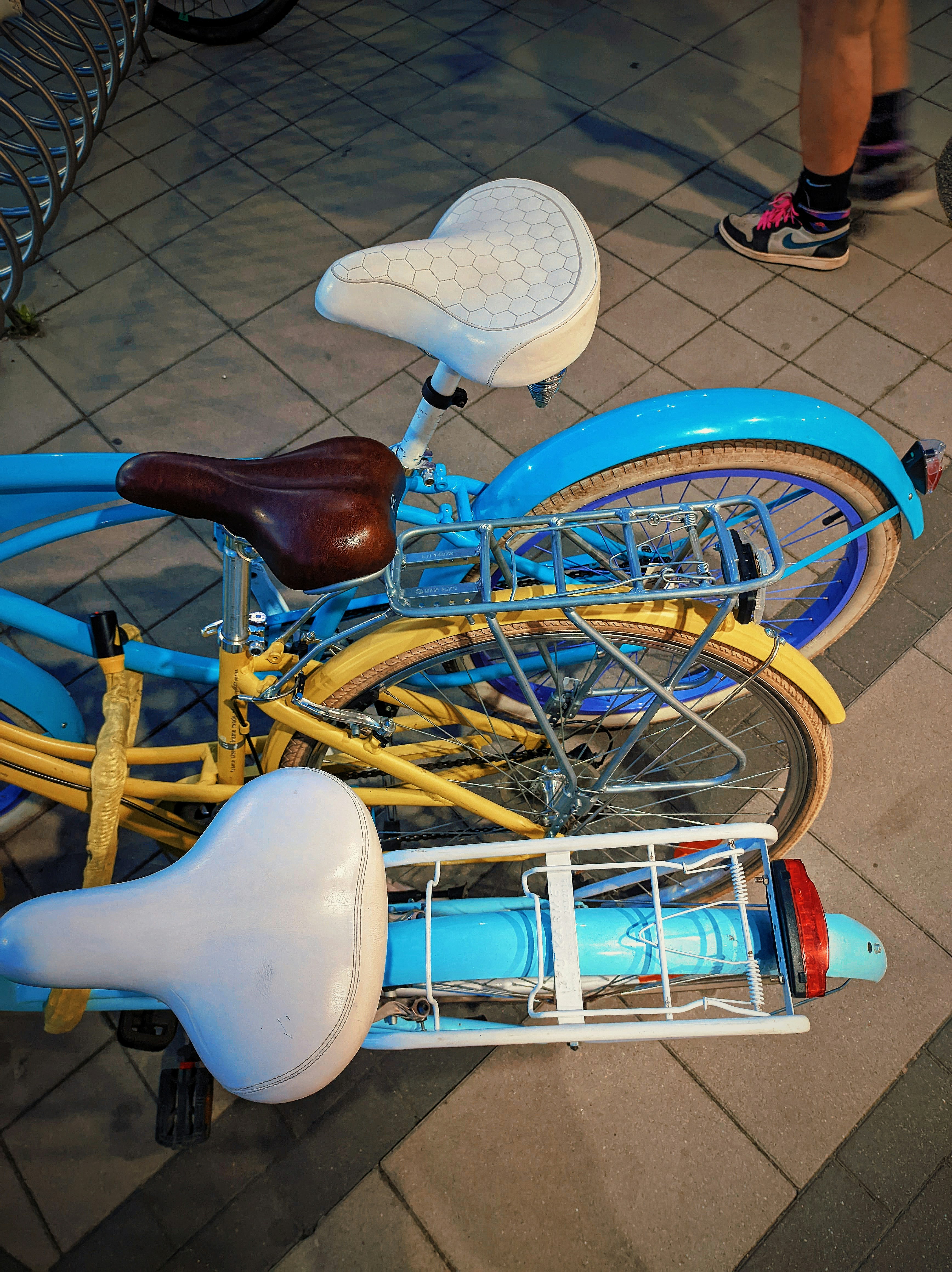 Stacked blue and yellow bicycles rest on a paved surface. White saddles and metal racks form a tangled urban vignette.