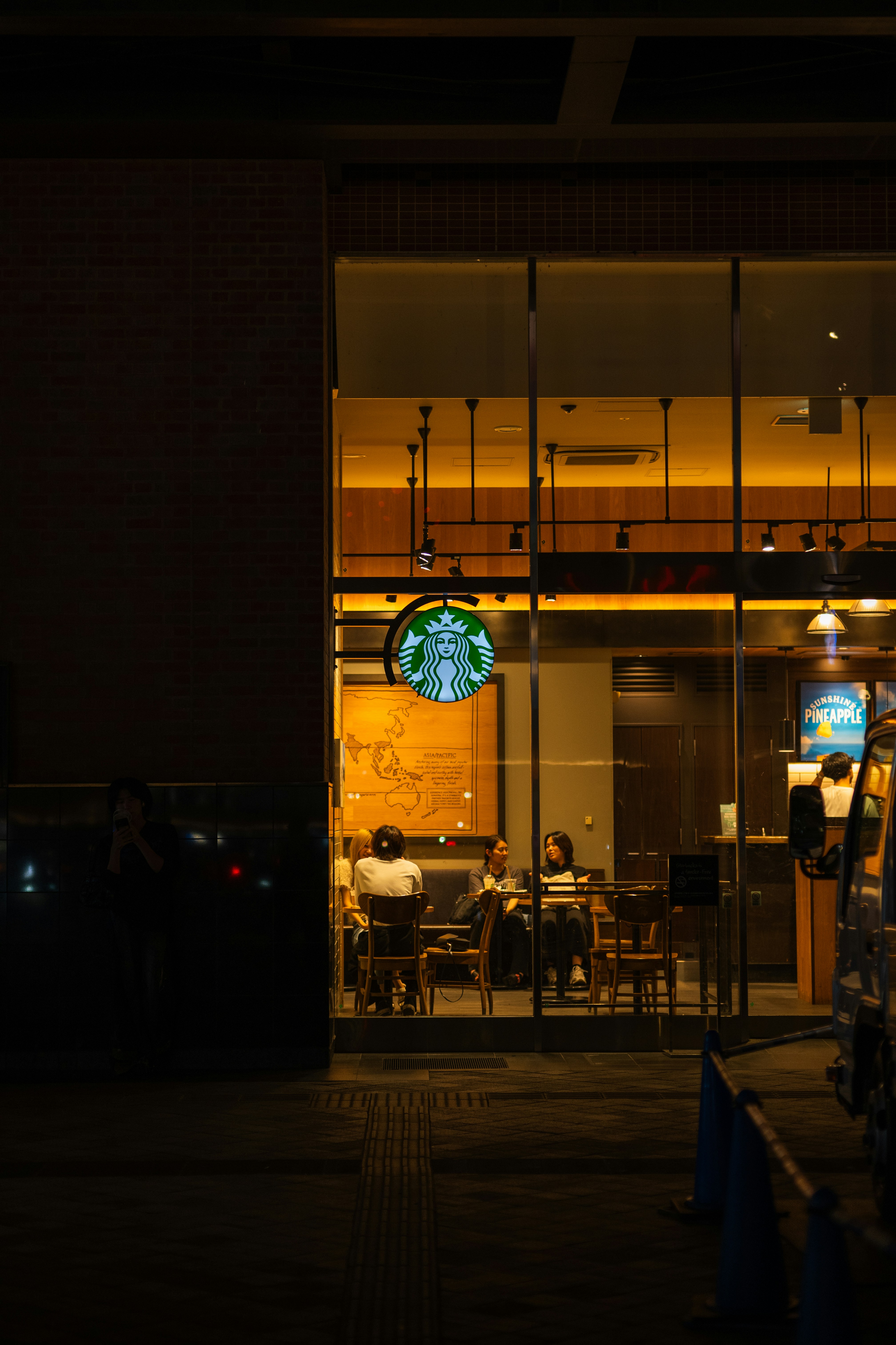 Warmly lit Starbucks interior visible through a glass storefront, with customers seated at tables and a glowing green siren logo.