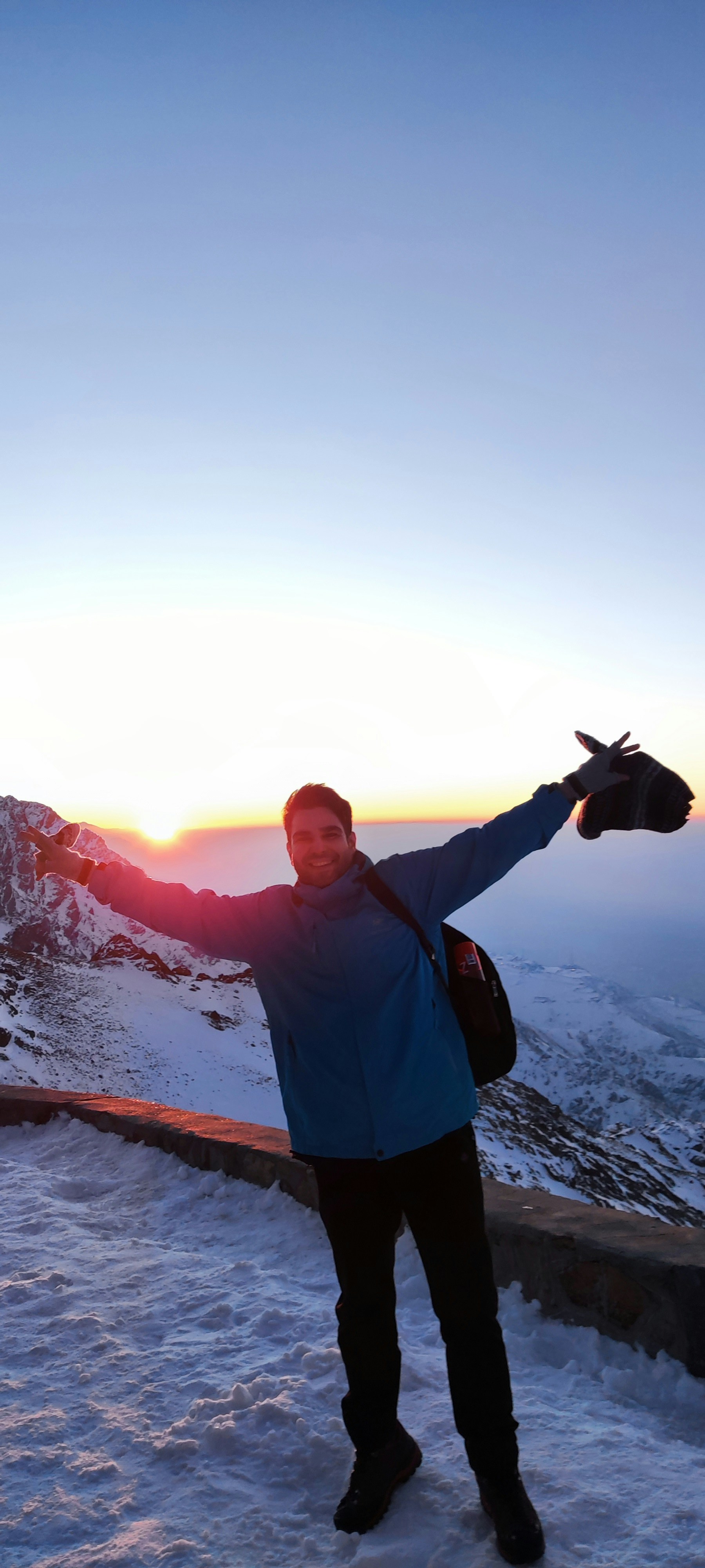 Photograph of a man in a blue jacket on a snow-covered ridge at sunrise, arms outstretched toward the sky.
