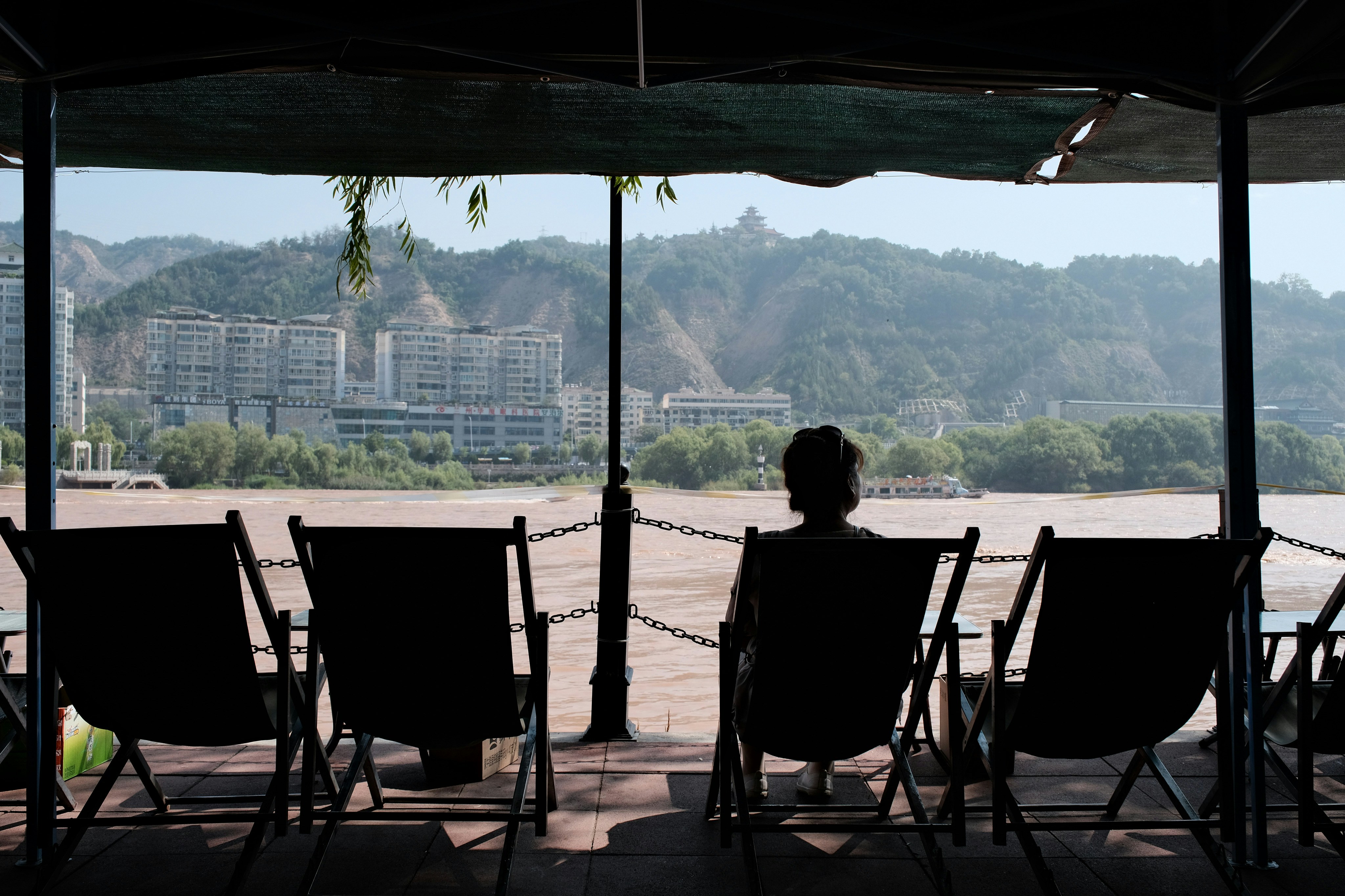 Silhouette of a person seated among empty chairs under a canopy, overlooking a river and distant buildings against a mountainous backdrop.