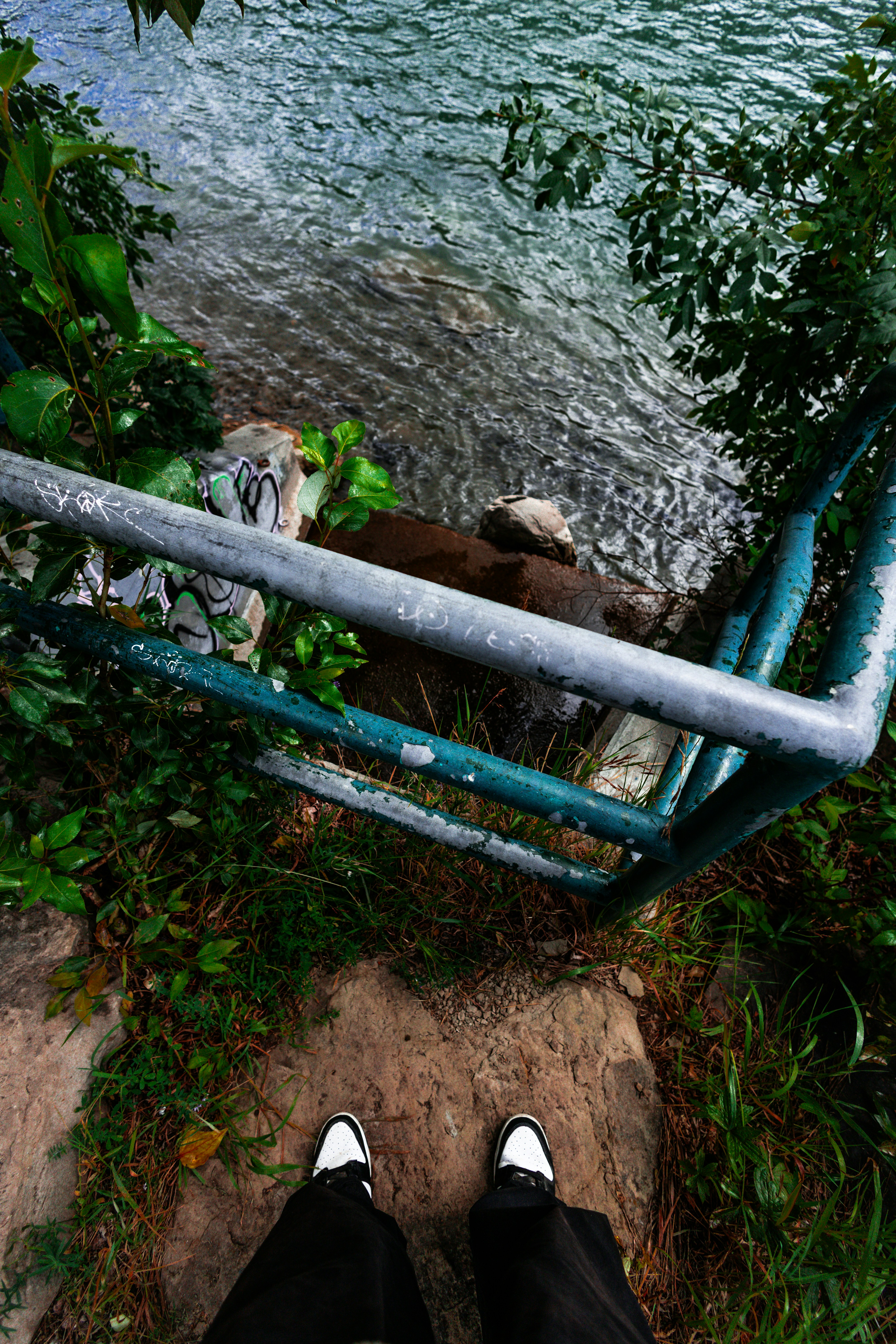 Footing at the River's Edge: sneakers on sunlit rock beside a graffiti-scarred railing and emerald water