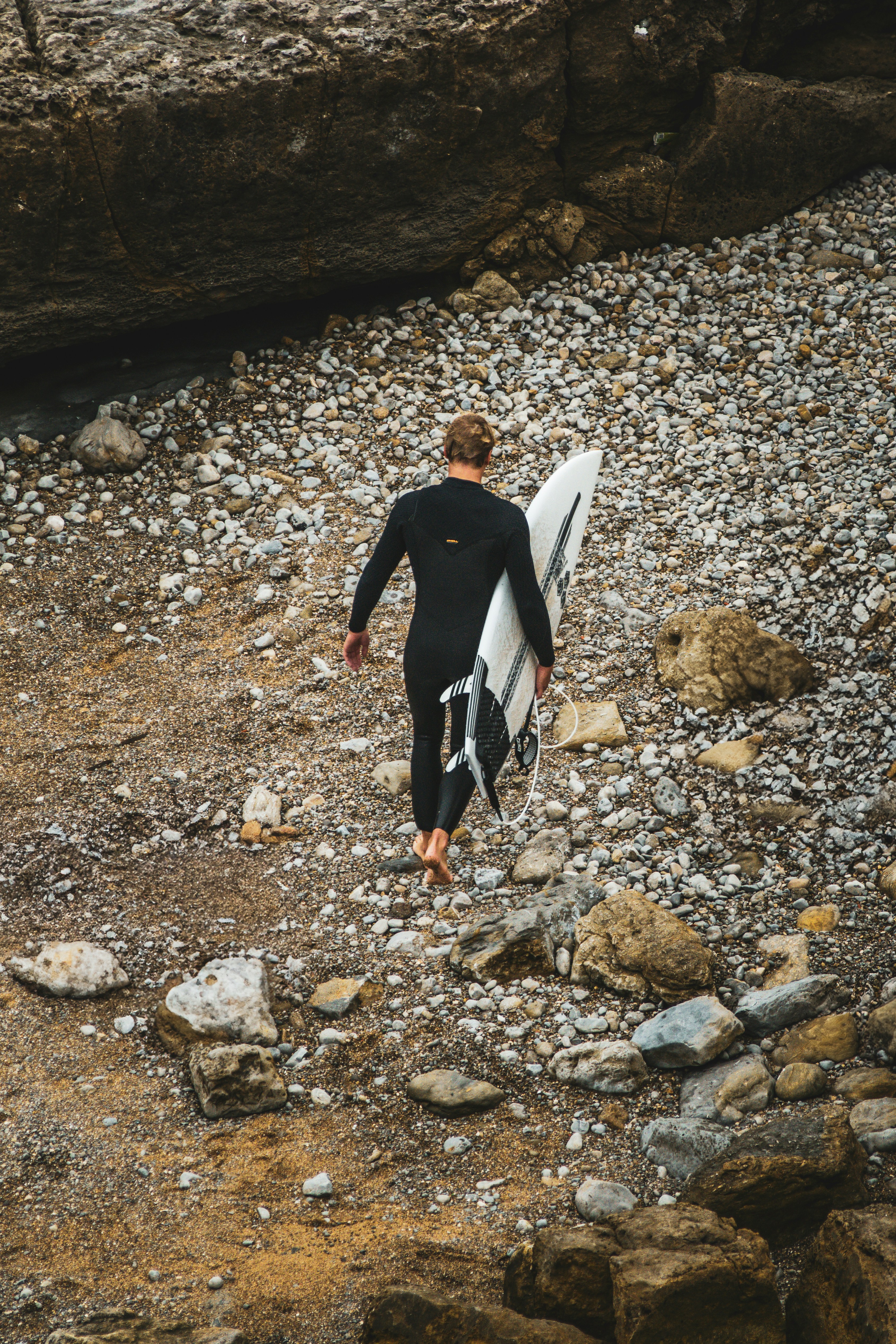 Shoreline Ascent: Surfer in Black Wetsuit Traverses Pebble Beach with Board