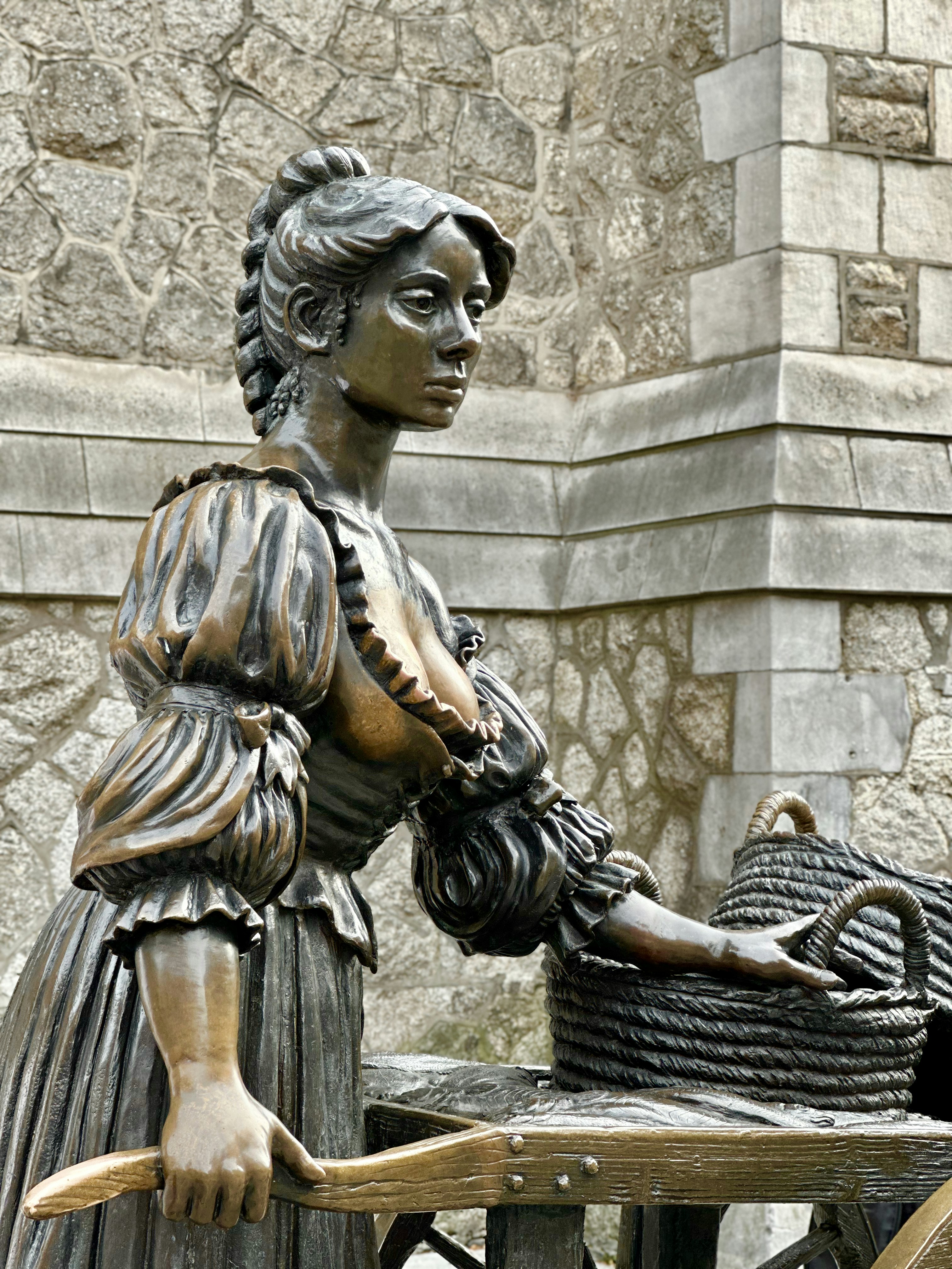 Bronze Statue of a Woman in Period Dress at a Wooden Workbench by a Stone Wall