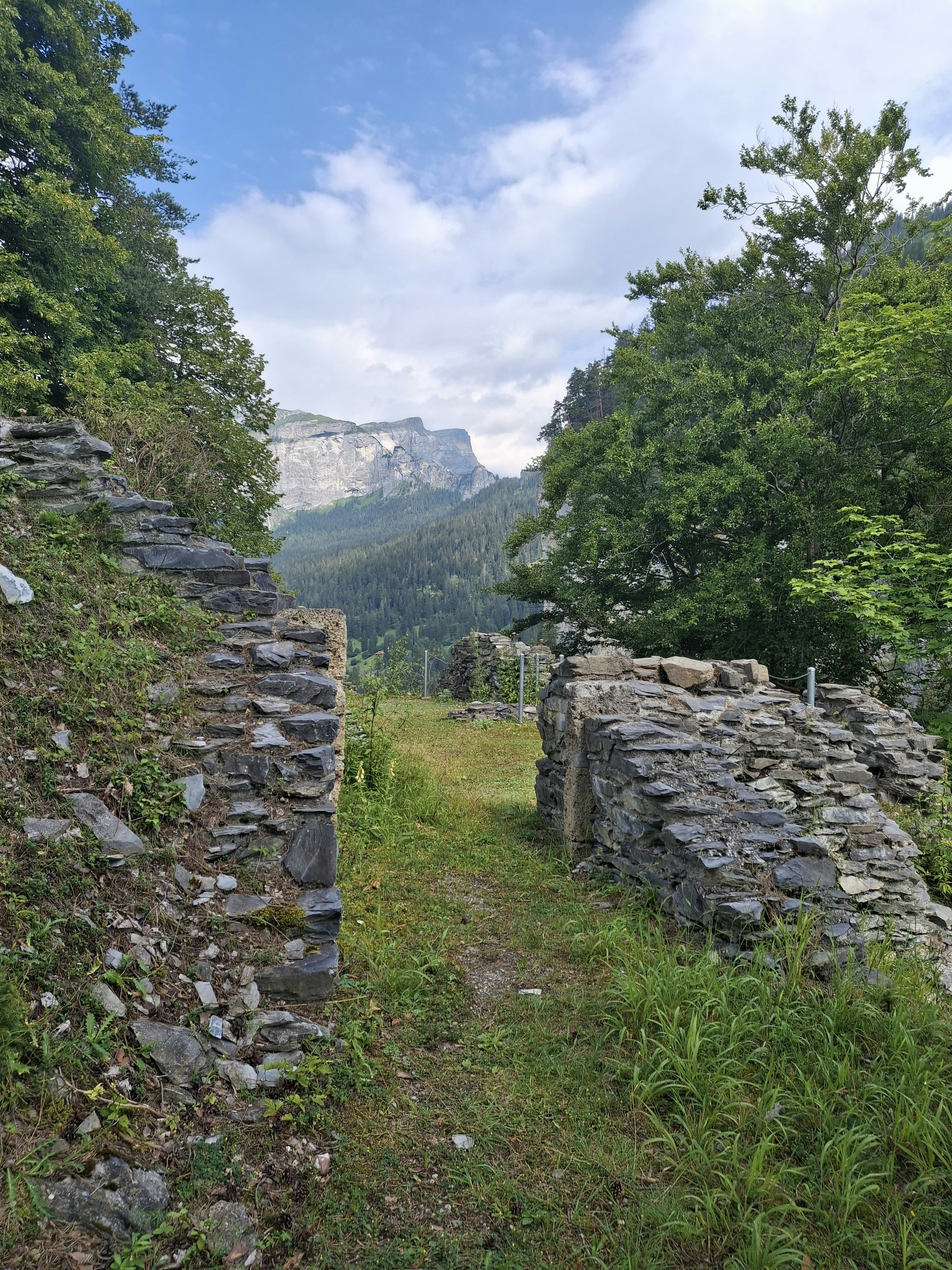 Narrow grassy path between weathered stone walls leads toward forested cliffs and a distant valley under a bright sky.