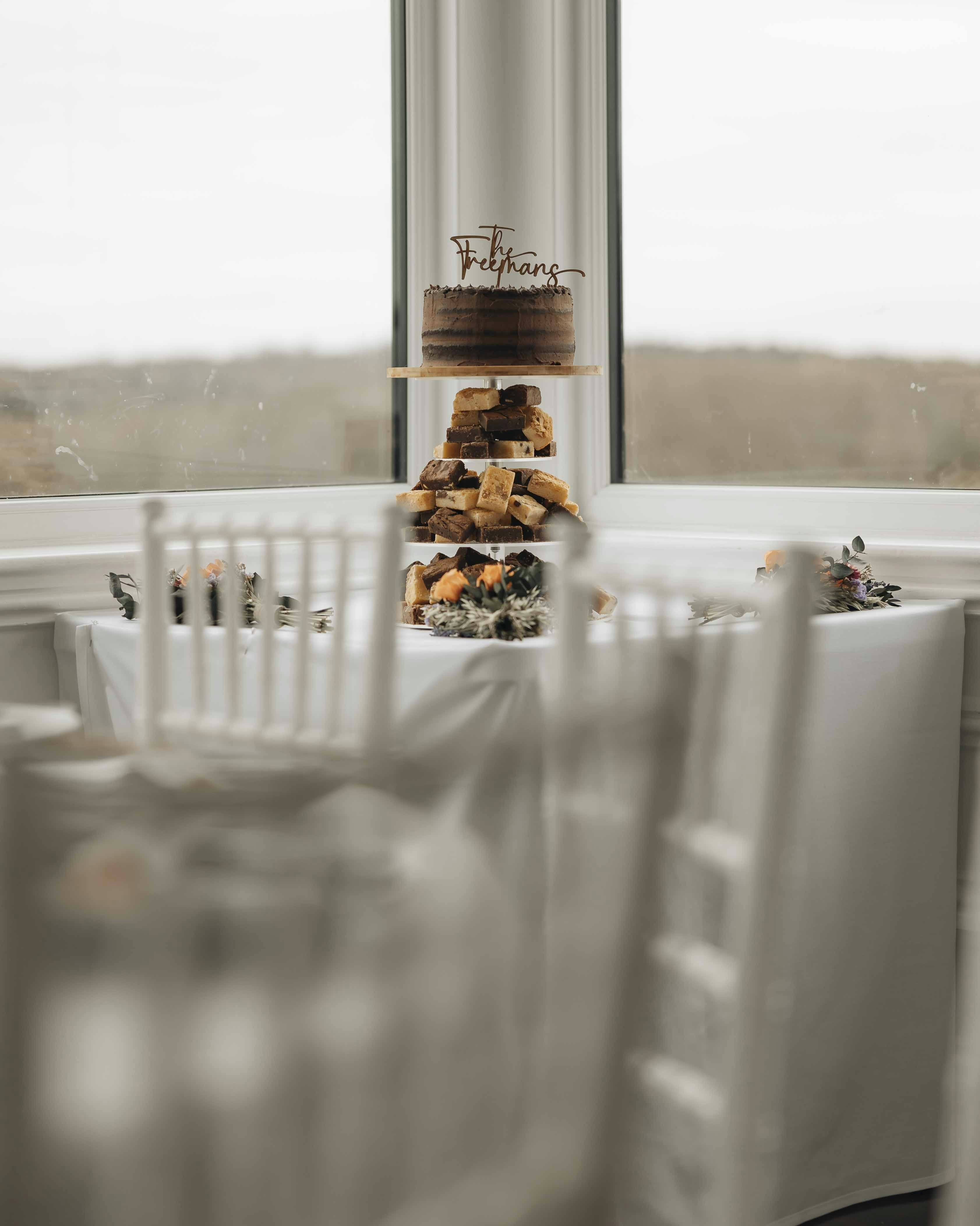 Wedding cake with bite-sized treats on a tiered stand near a window, surrounded by soft-focus chairs.