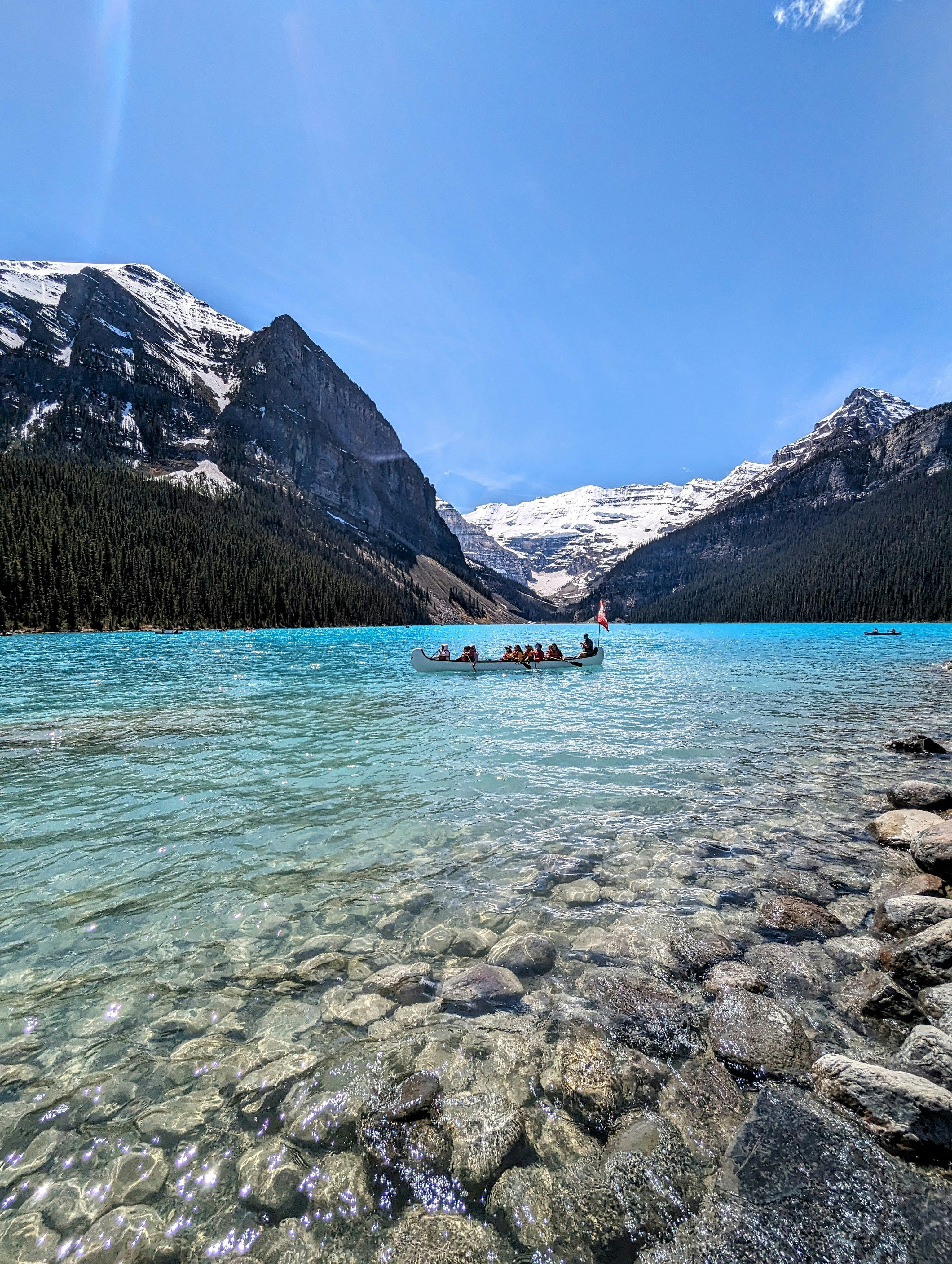 Turquoise glacier-fed lake framed by snow-dusted mountains and evergreen forests, with a small boat carrying passengers gliding near the center. Clear, sunny conditions enhance color and depth of the scene.