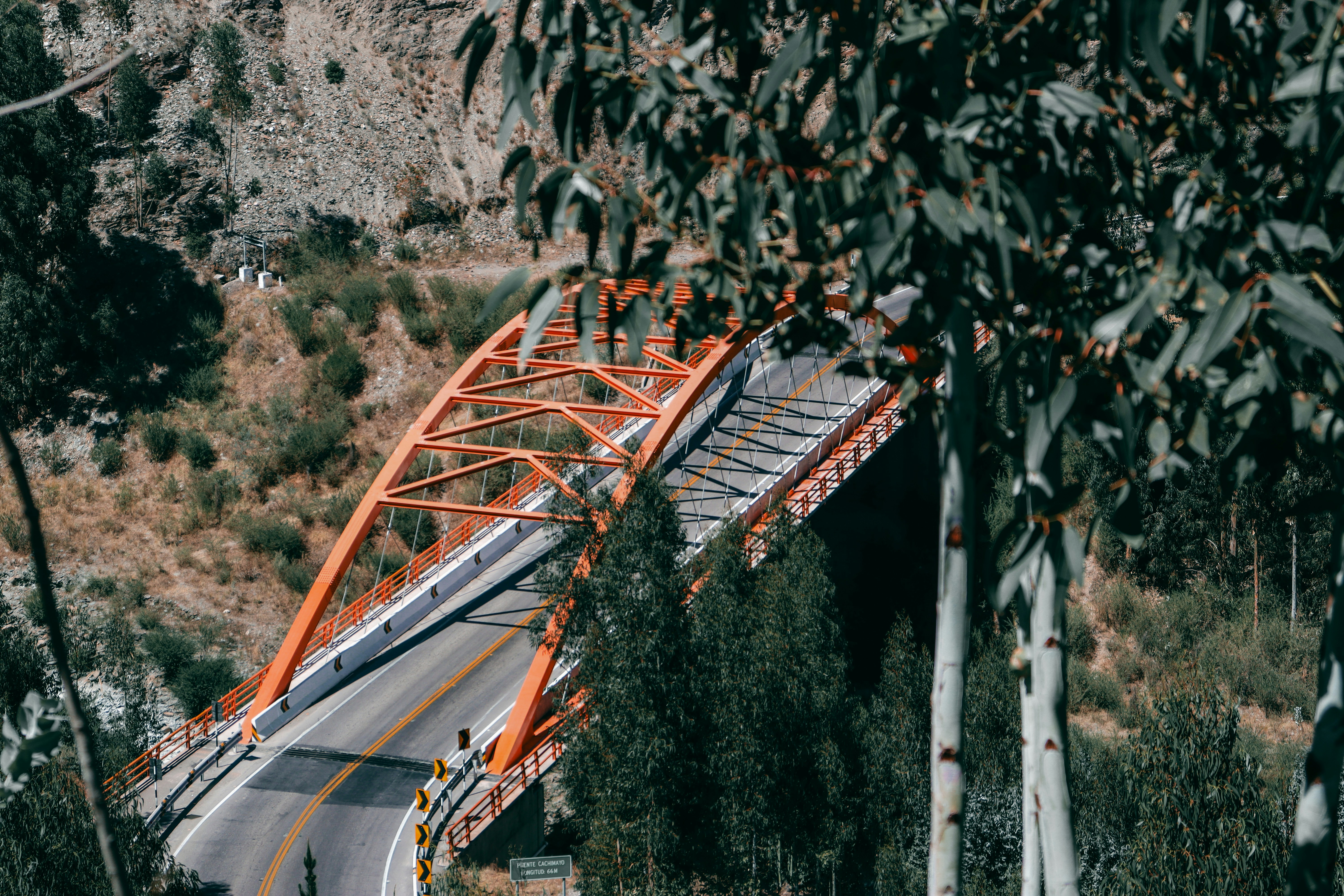 Orange bridge arches over a green valley, framed by trees and rugged terrain.