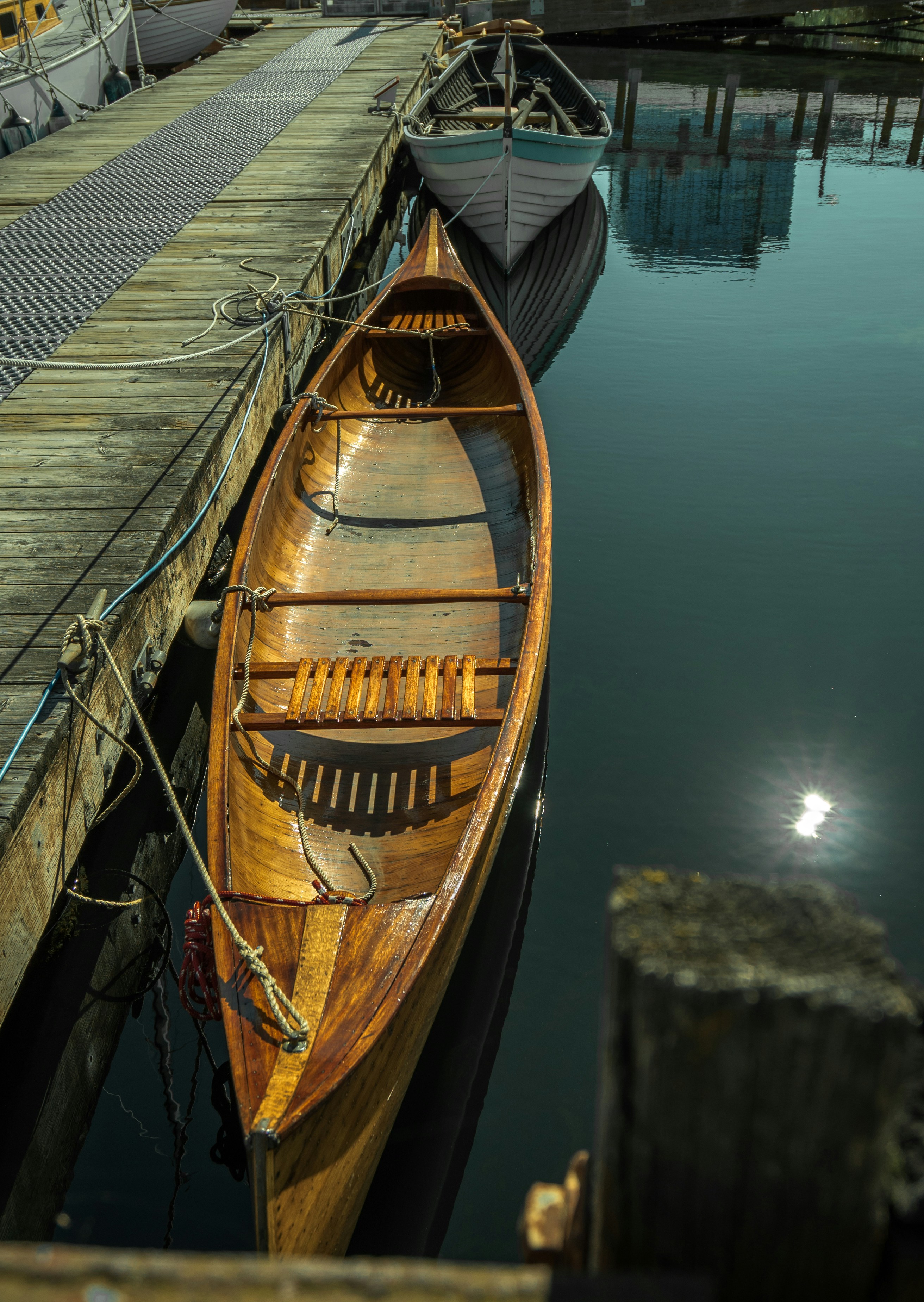 A beautifully crafted wooden canoe rests beside a dock, its reflection shimmering in the calm water. Sunlight glints off the surface, enhancing the tranquil atmosphere.