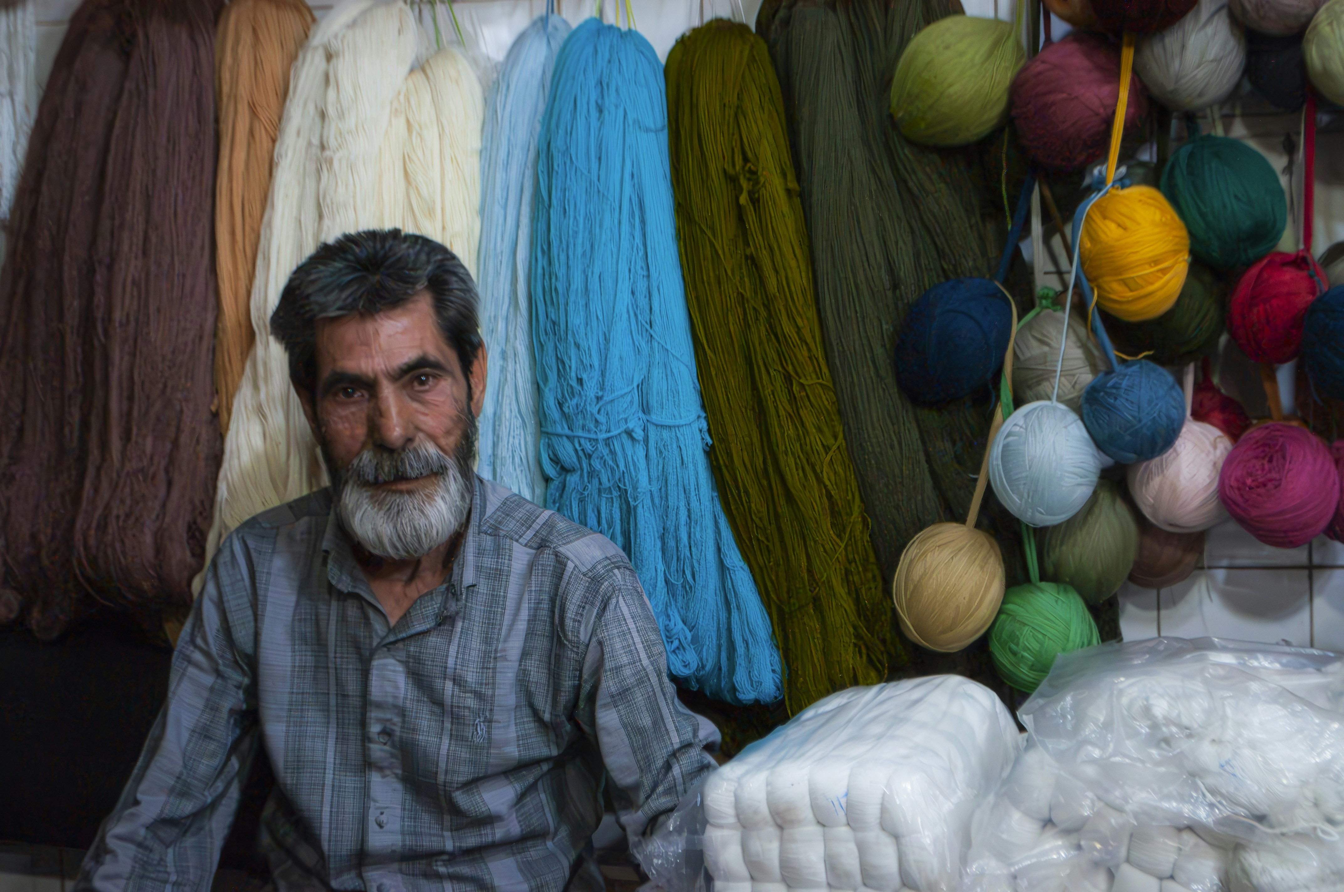 Man seated in a yarn shop surrounded by colorful skeins and balls of yarn.