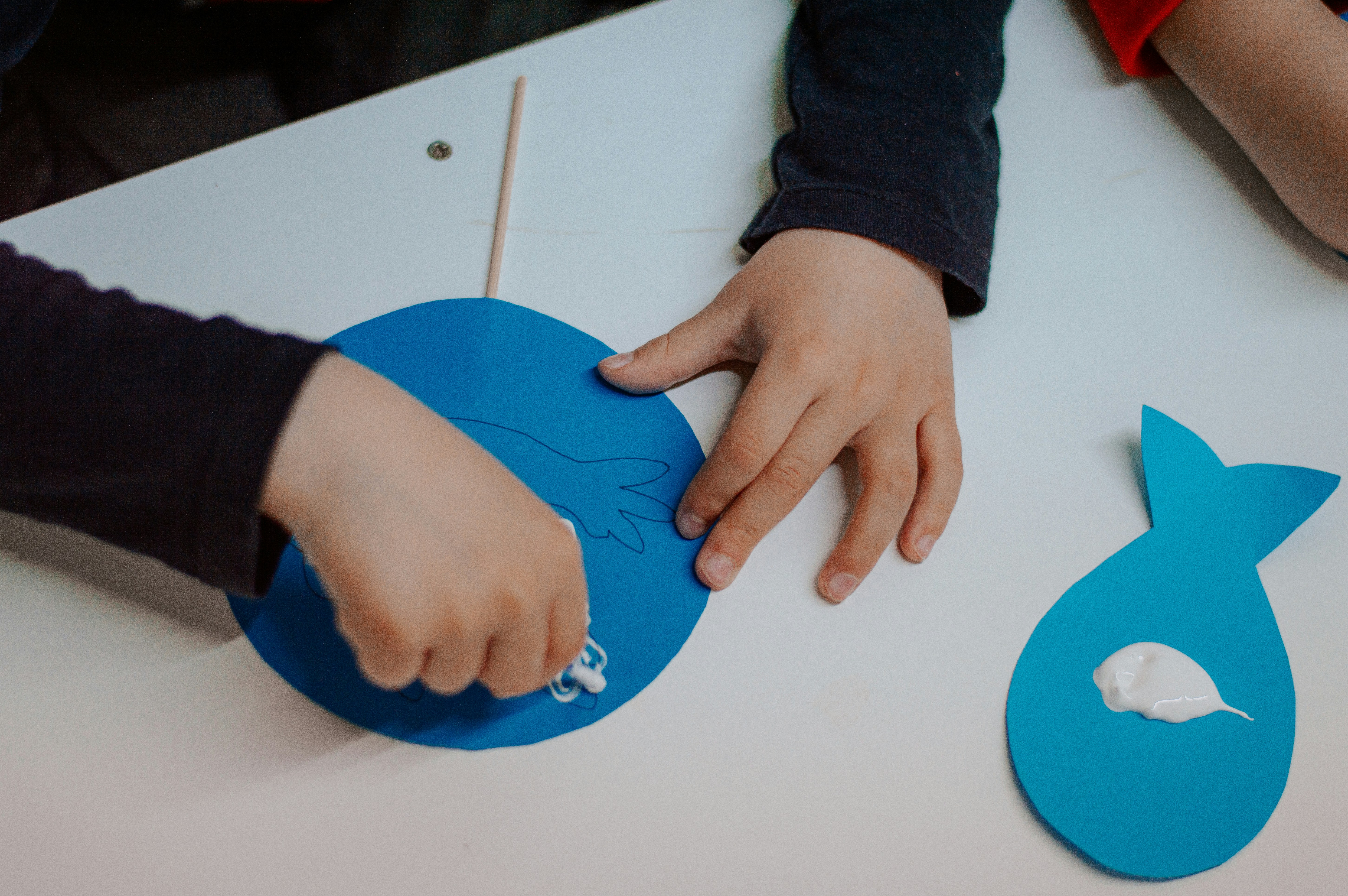 Children's hands assembling paper cutouts of a blue whale on a white table.