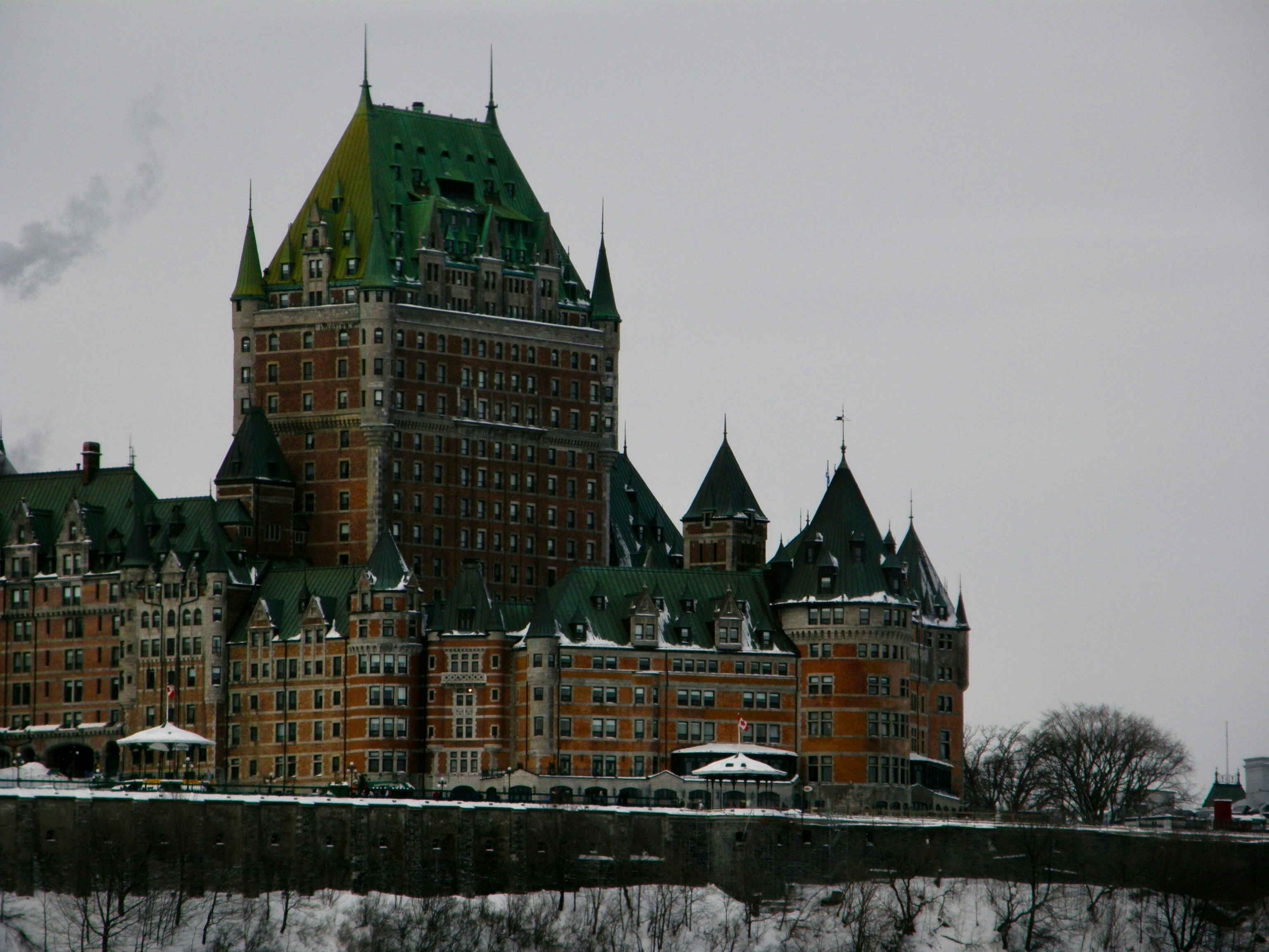 Château Frontenac dominates a snow-dusted riverfront, its green copper roofs and brick towers forming a fortresslike silhouette. An overcast sky and bare trees amplify the winter mood.