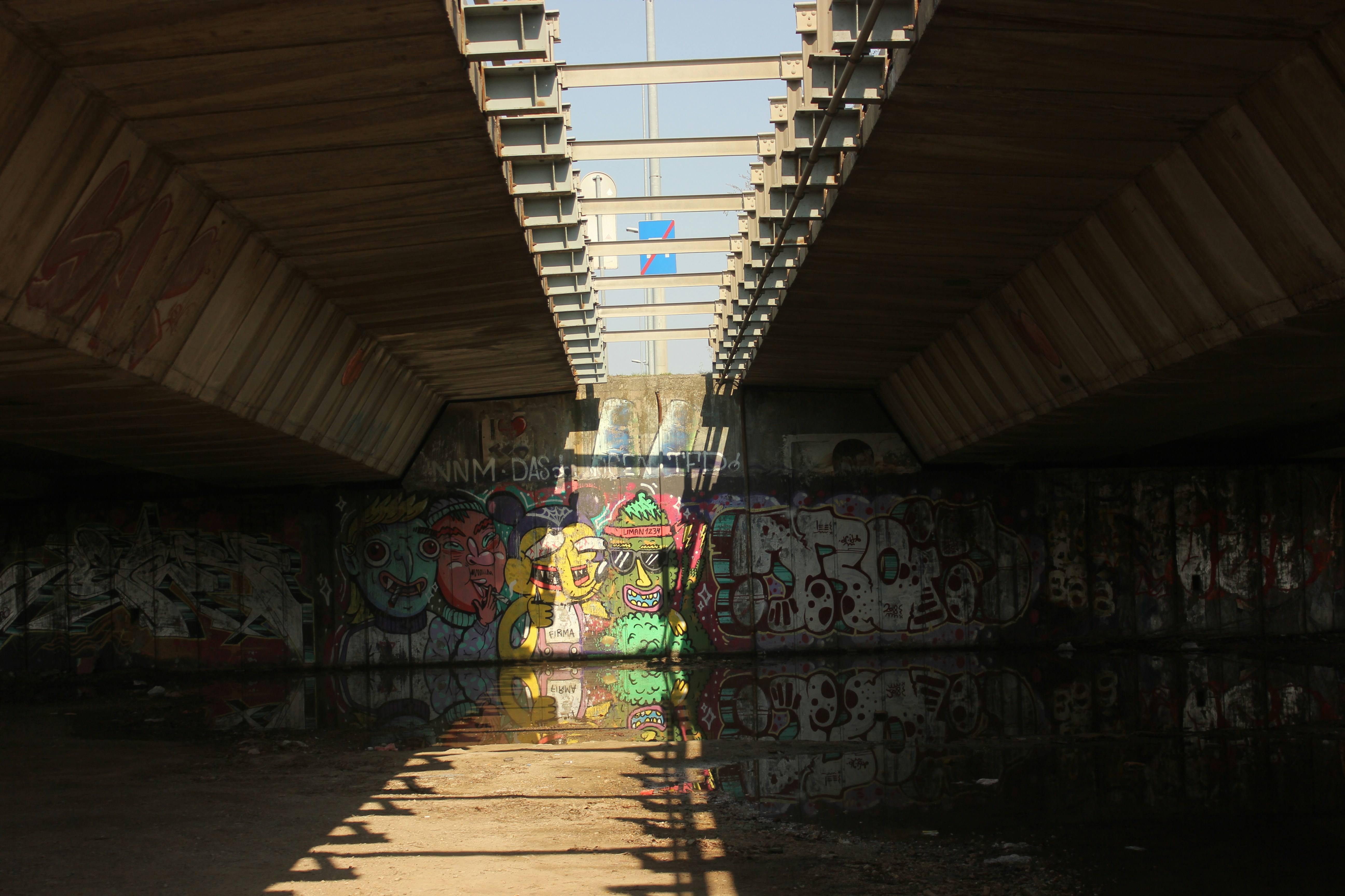 Graffiti-covered walls under a bridge with reflected light on a shallow pool of water.