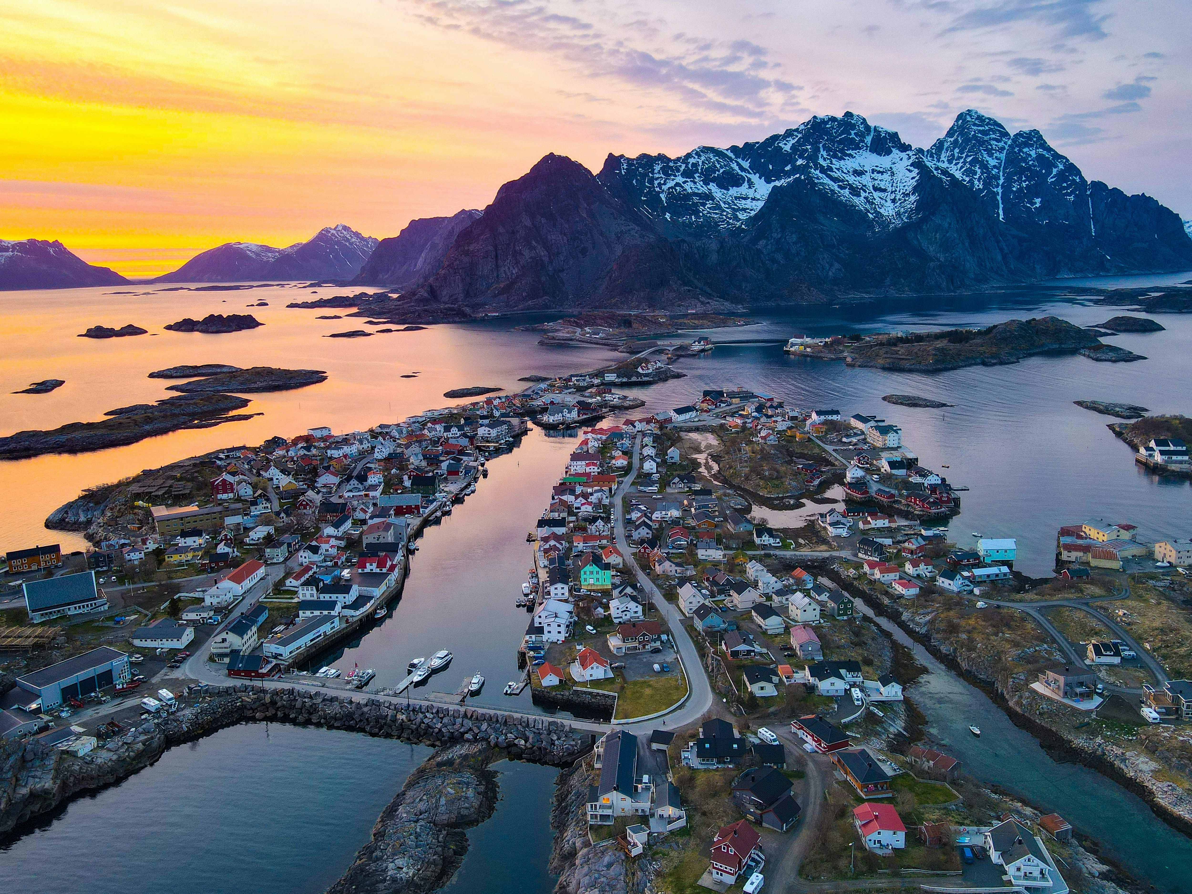 Coastal village nestled between calm waters and rugged snow-capped mountains under a vibrant sunset sky.